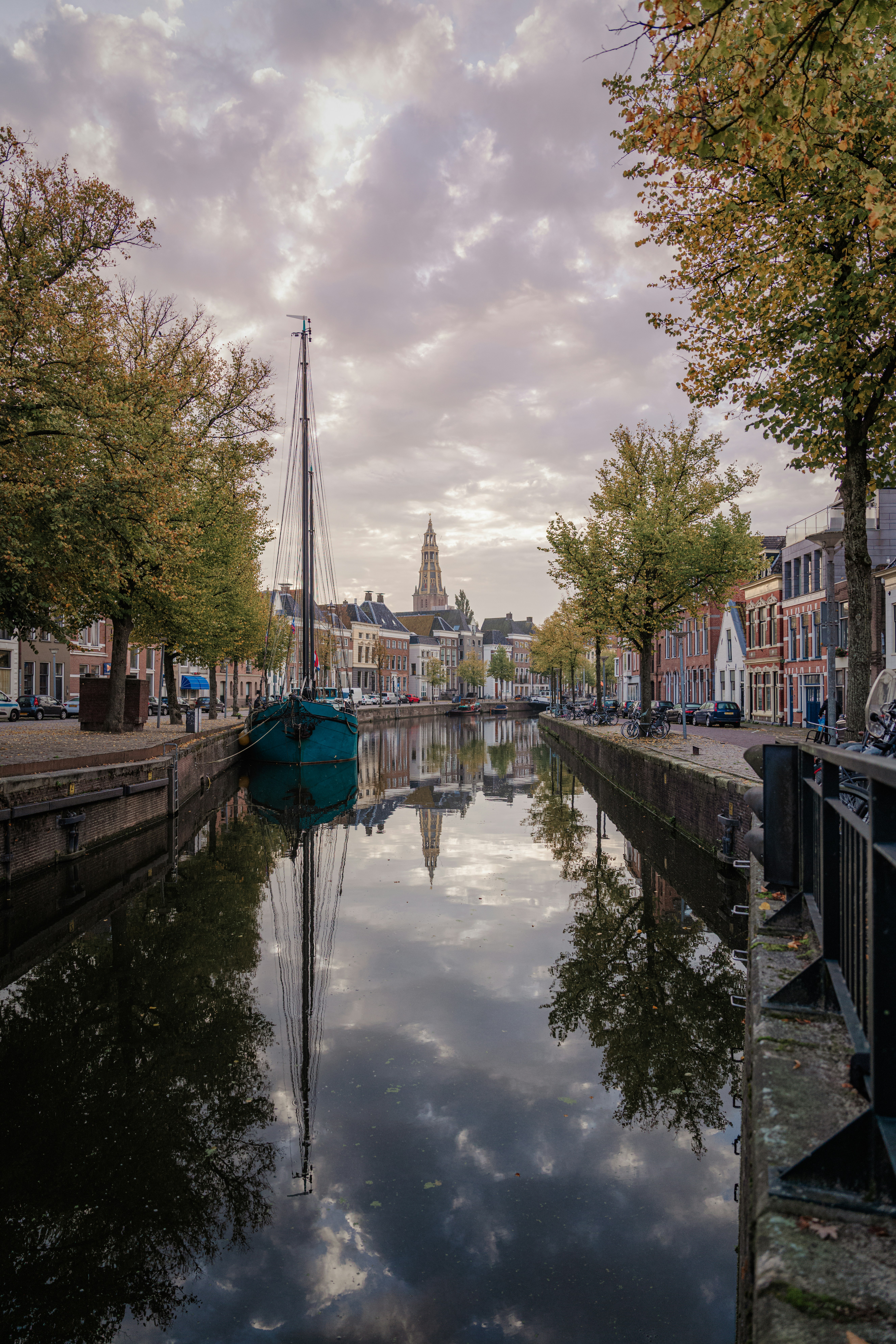 A serene canal scene featuring a sailboat and tree-lined banks, reflecting the charming architecture and cloudy sky above.