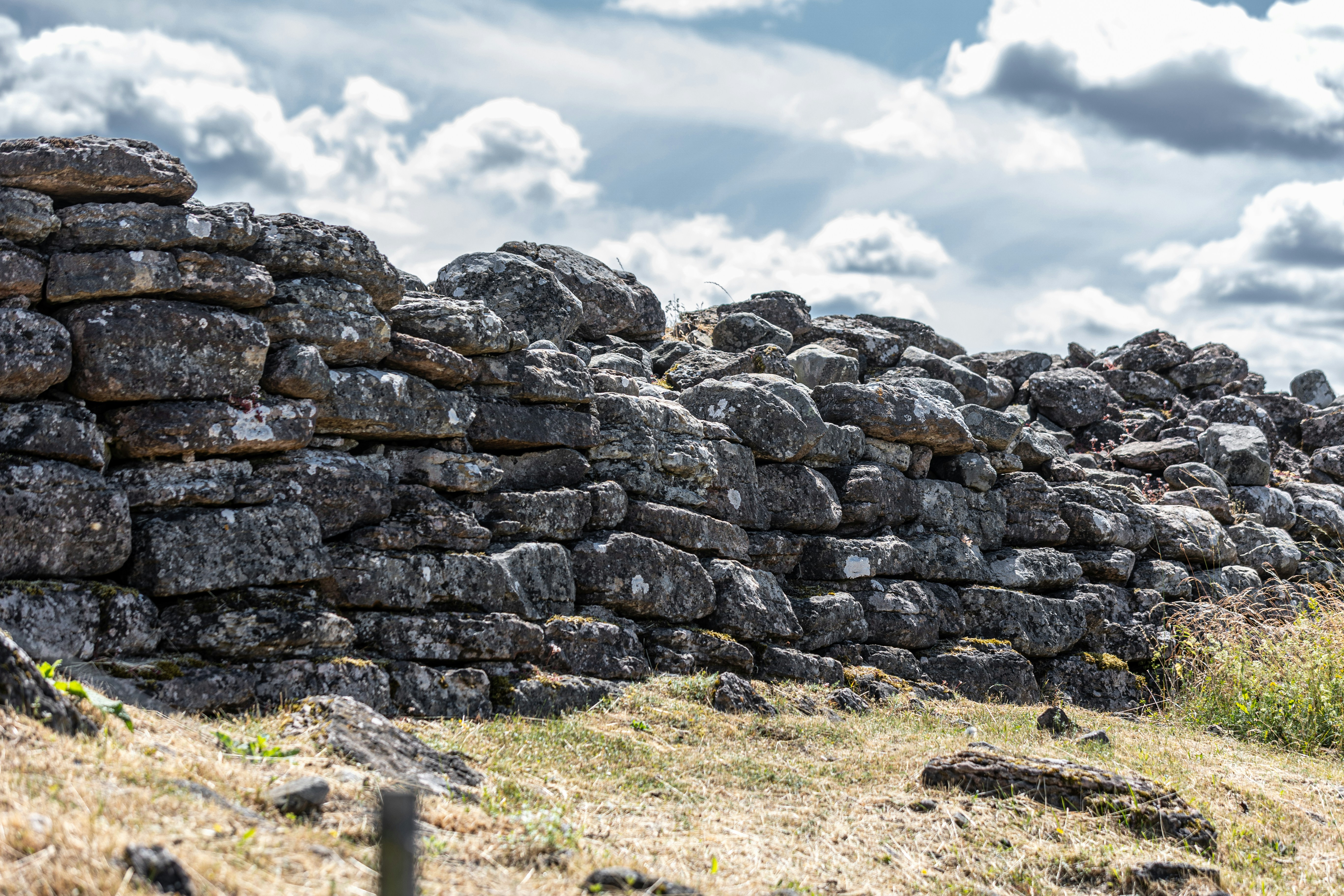 Weathered stone wall remnants display intricate textures against a backdrop of dynamic clouds. Nature reclaims the land around the historic structure.
