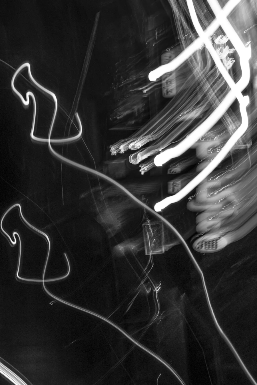 Black-and-white long-exposure scene with bright light trails sweeping across a dark frame, creating abstract curves and streaks. Blurred shapes and highlights cluster on the right side.