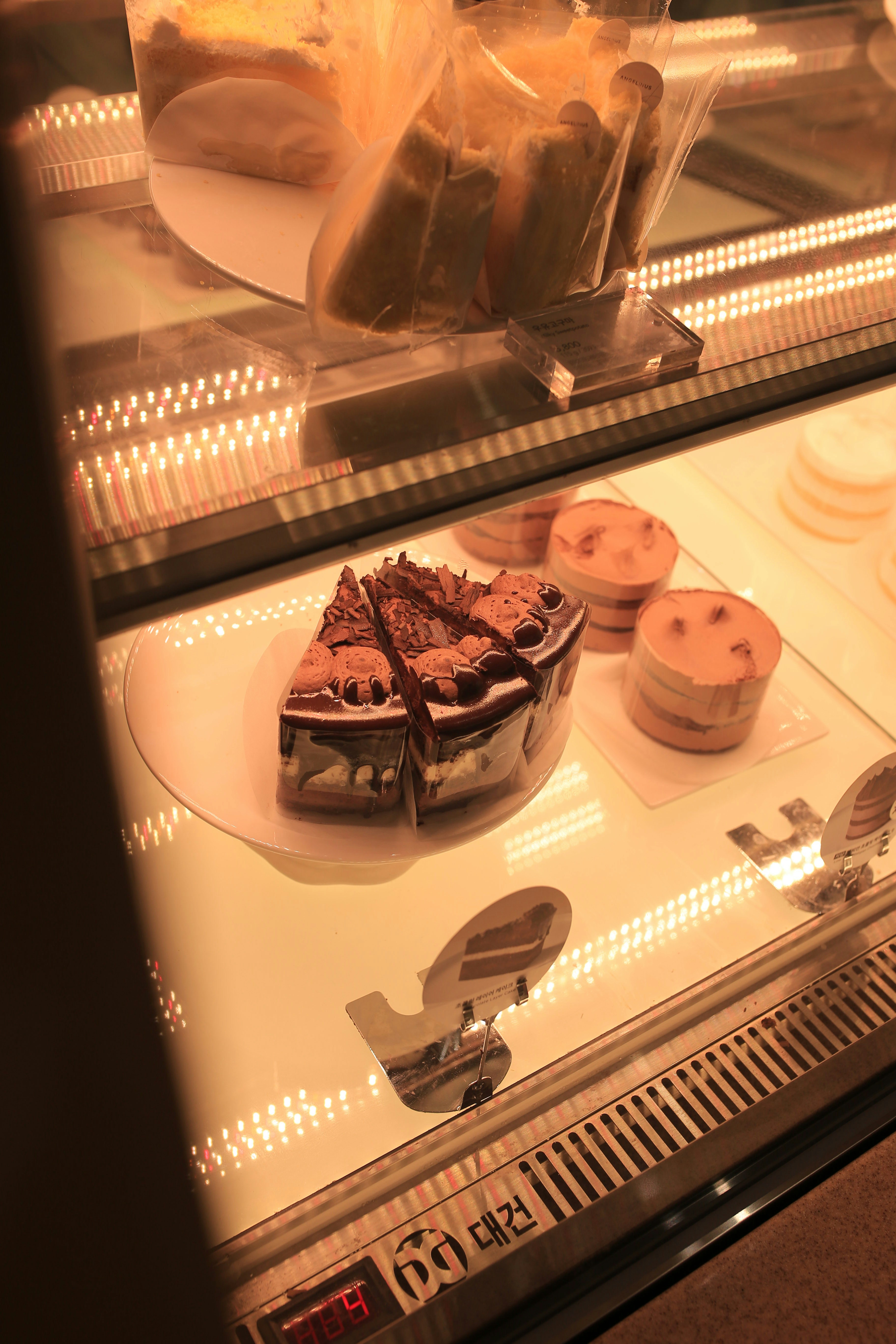 Chocolate cake slice elegantly displayed in a dessert case, surrounded by other tempting treats. The warm lighting enhances the inviting atmosphere.