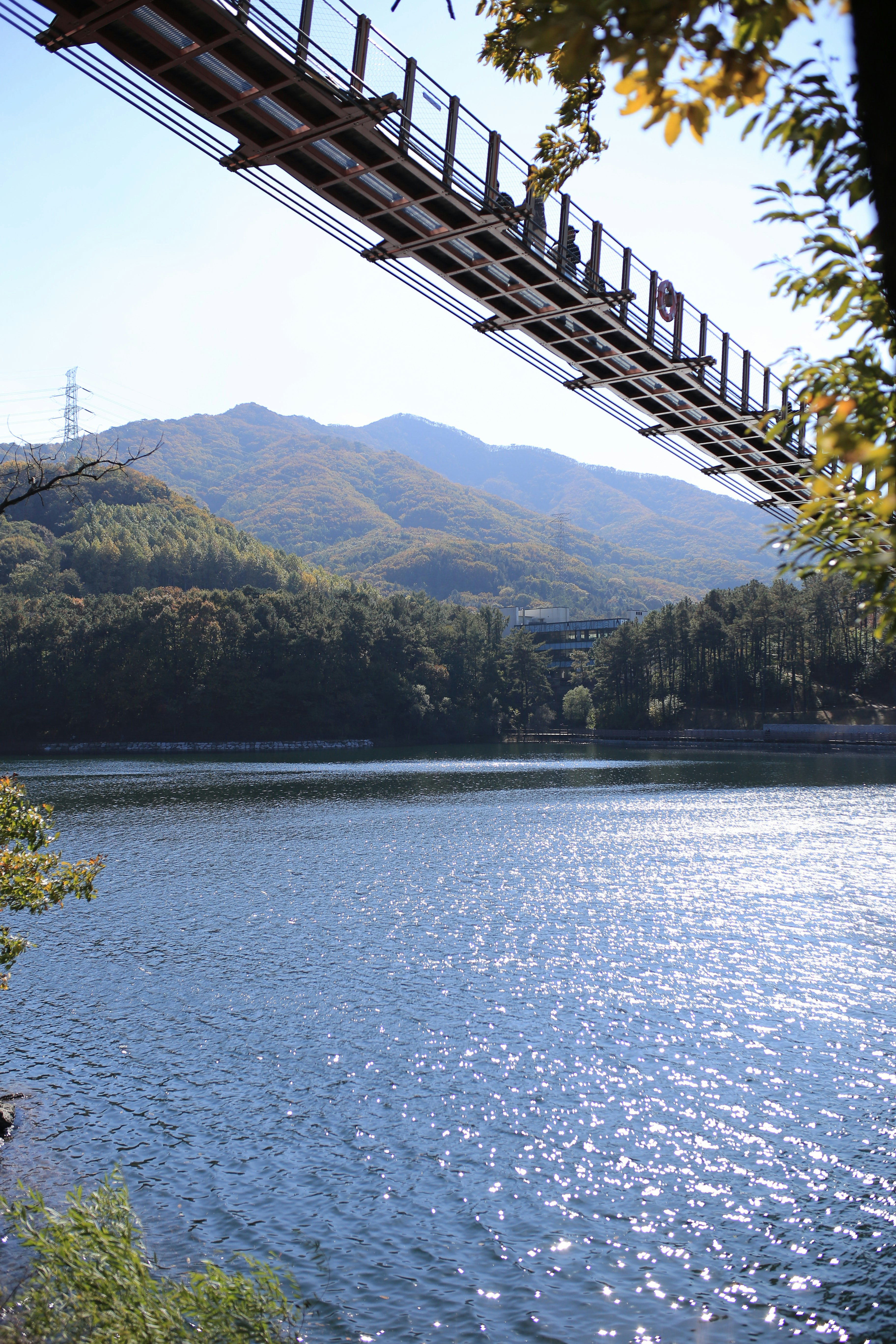 Suspension bridge arching over a calm lake, framed by autumn foliage and mountains in the background.