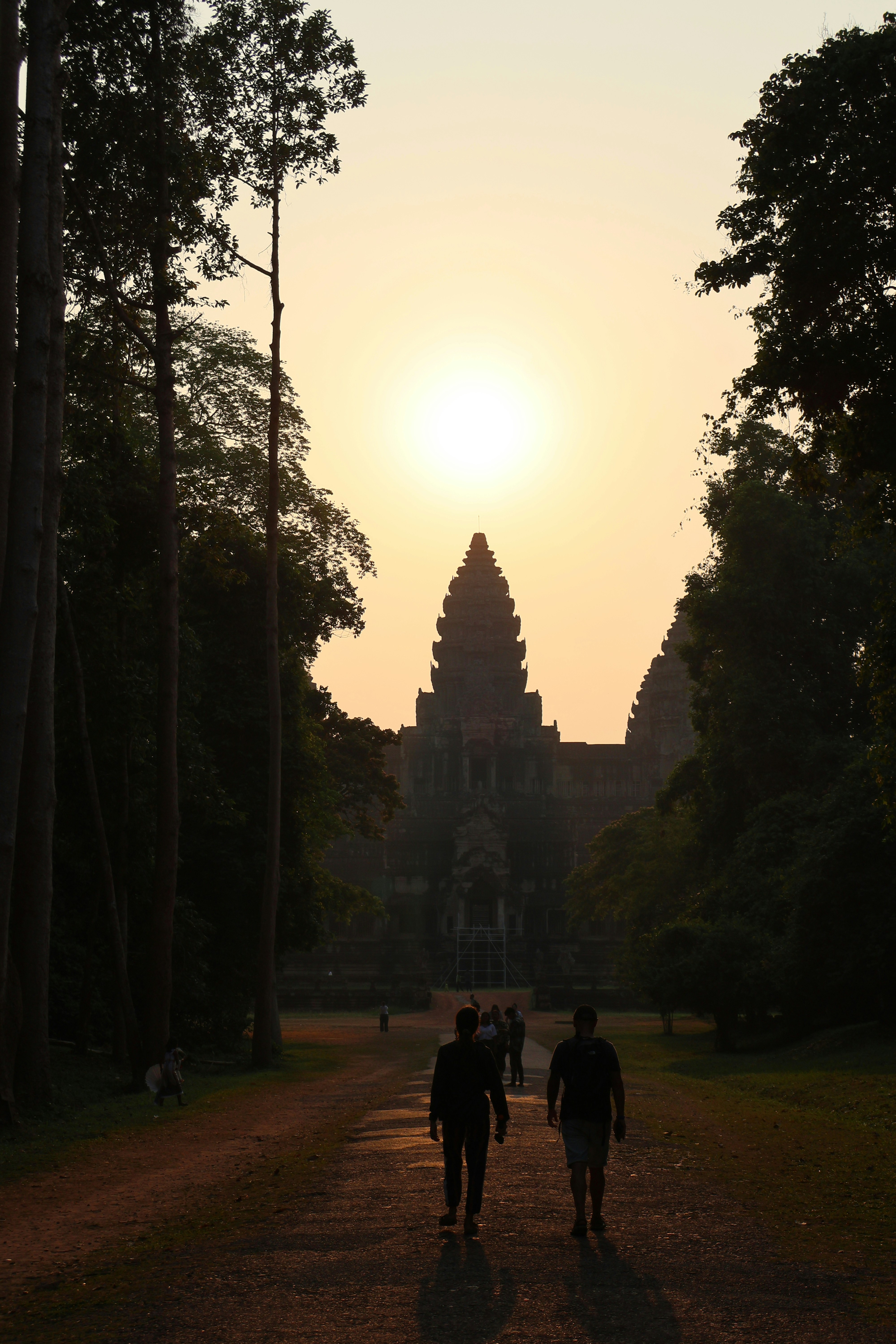 Silhouetted figures walk along a pathway leading to the majestic Angkor Wat temple, framed by towering trees and a glowing sunrise.