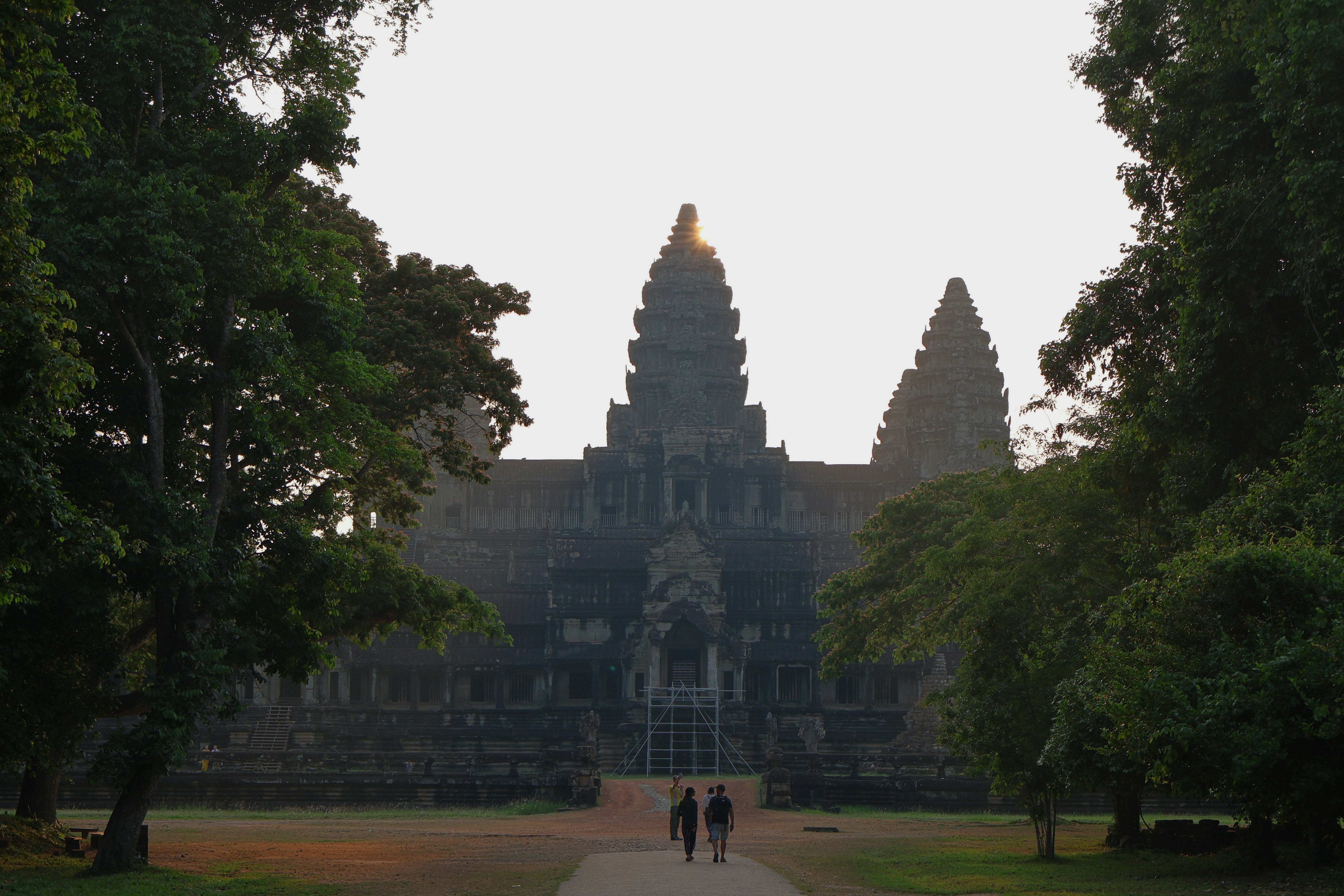 Silhouetted figures walking towards the grand Angkor Wat temple, framed by lush greenery and a soft morning light.