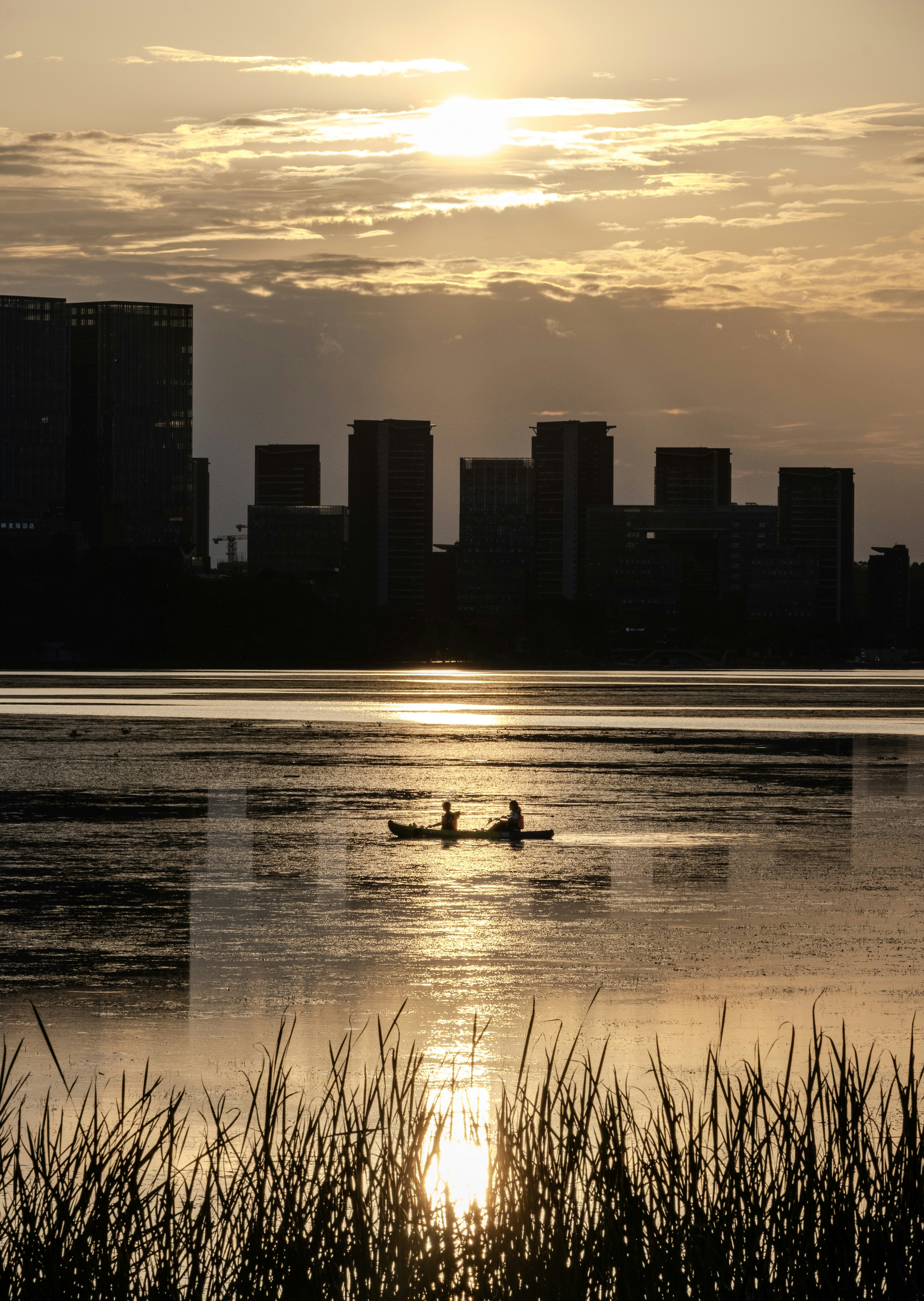 Two kayakers glide across a tranquil water surface, framed by silhouetted city buildings under a golden sunset. The scene captures a moment of peace amidst urban life.
