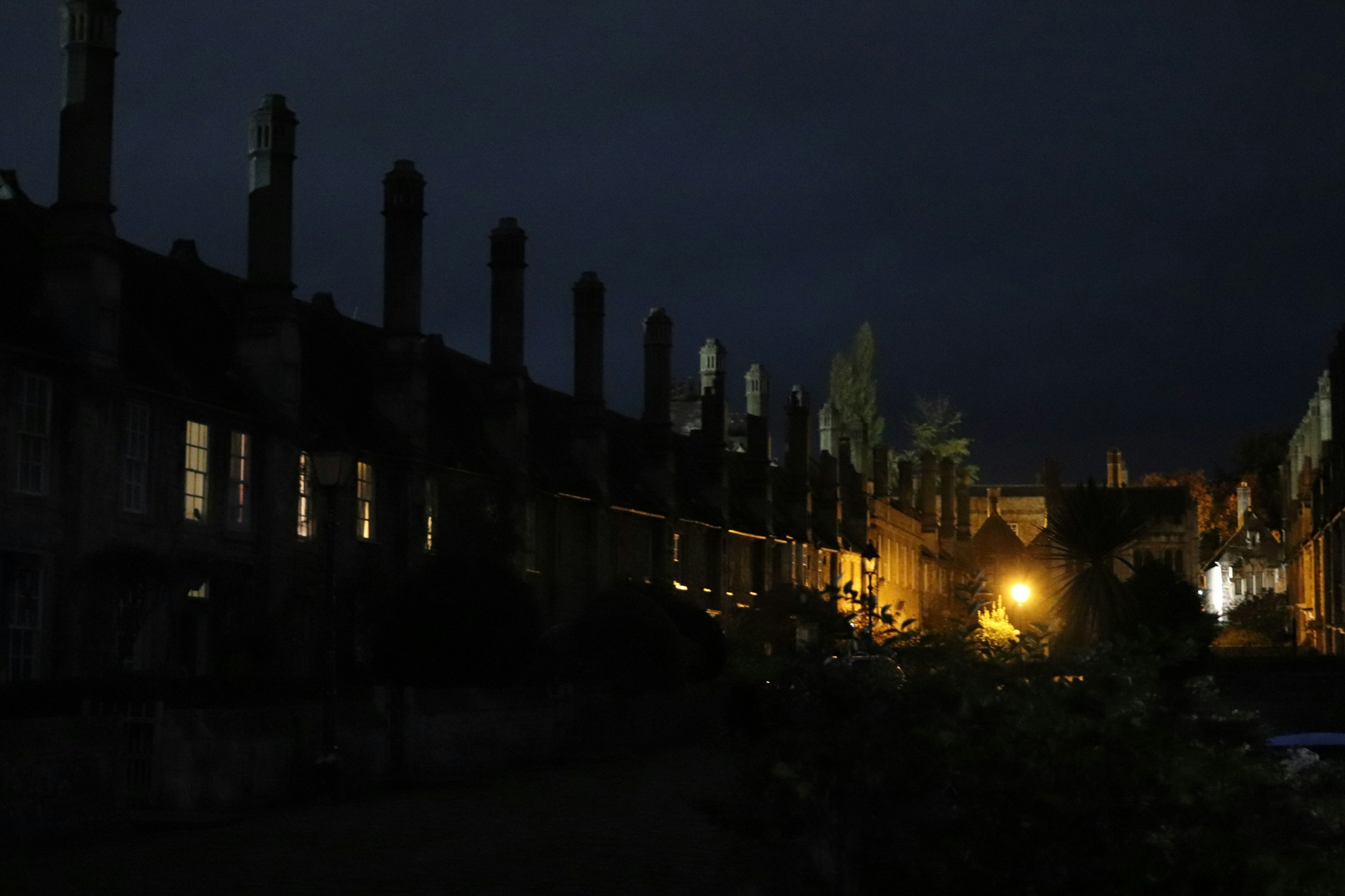 Historic buildings line a dimly lit street under a night sky, with warm light spilling from windows and a distant streetlamp illuminating the scene.