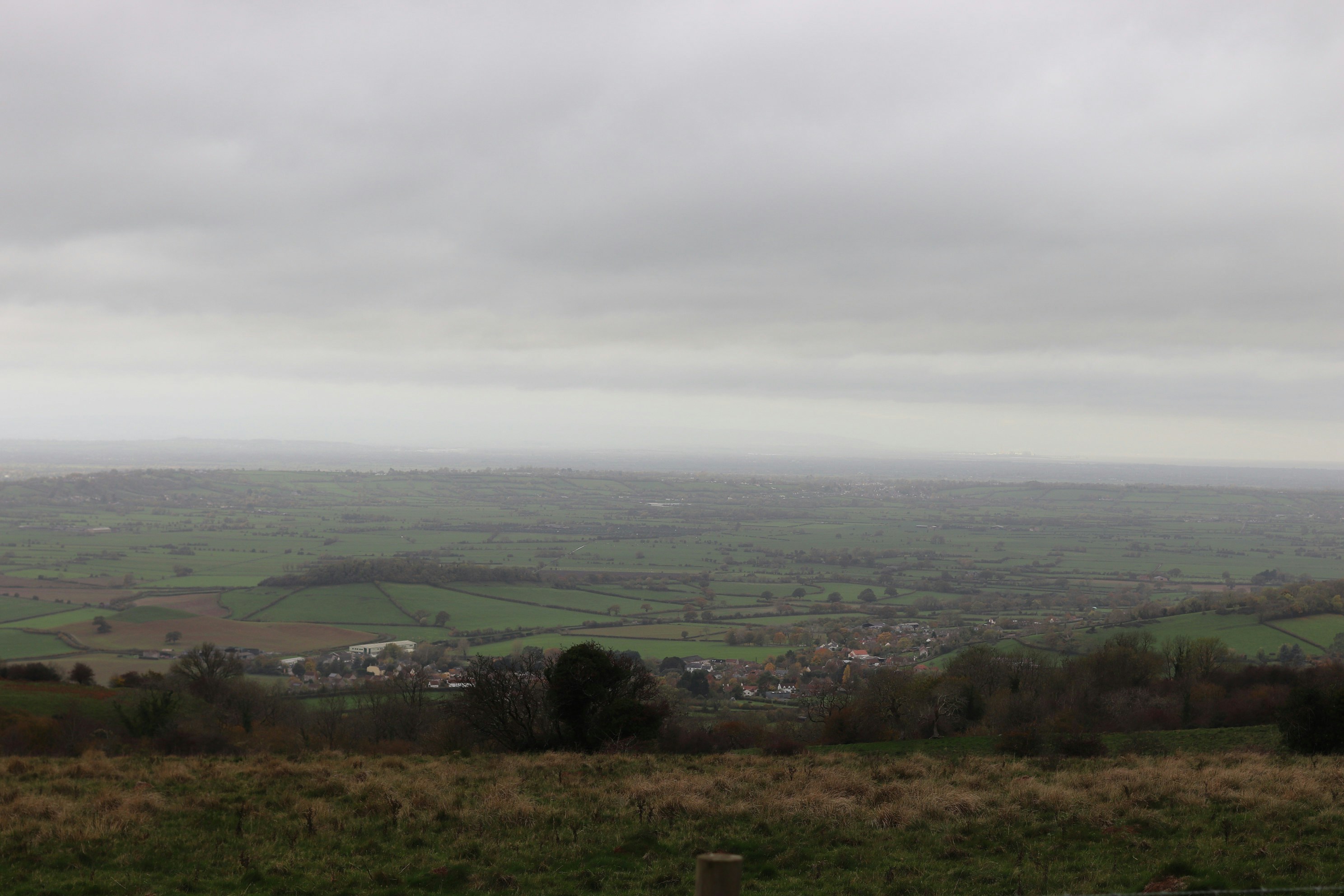 Expansive view of rolling green fields and a distant village under a cloudy sky, capturing the serene beauty of rural England.