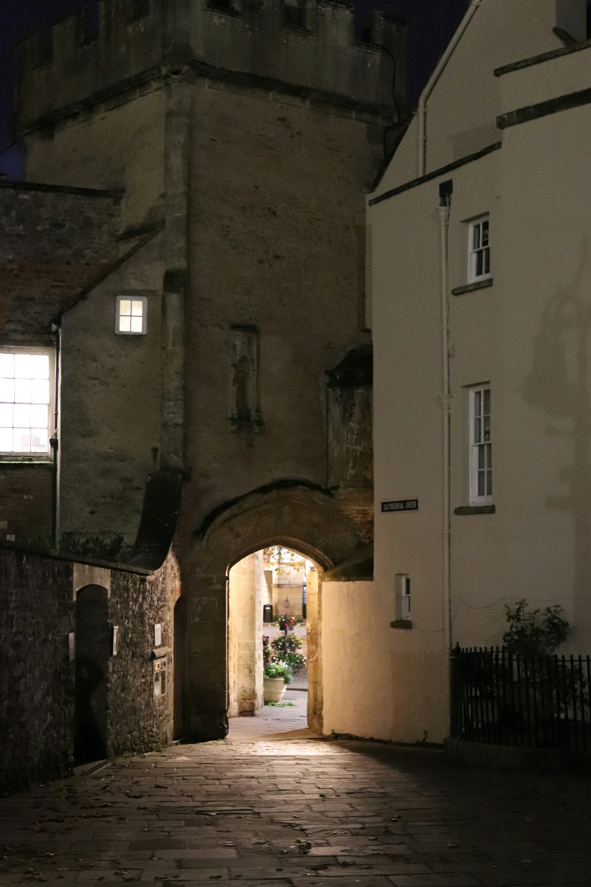 Illuminated archway leading to a quaint courtyard, framed by historic stone walls and a softly lit window. A serene atmosphere invites exploration.