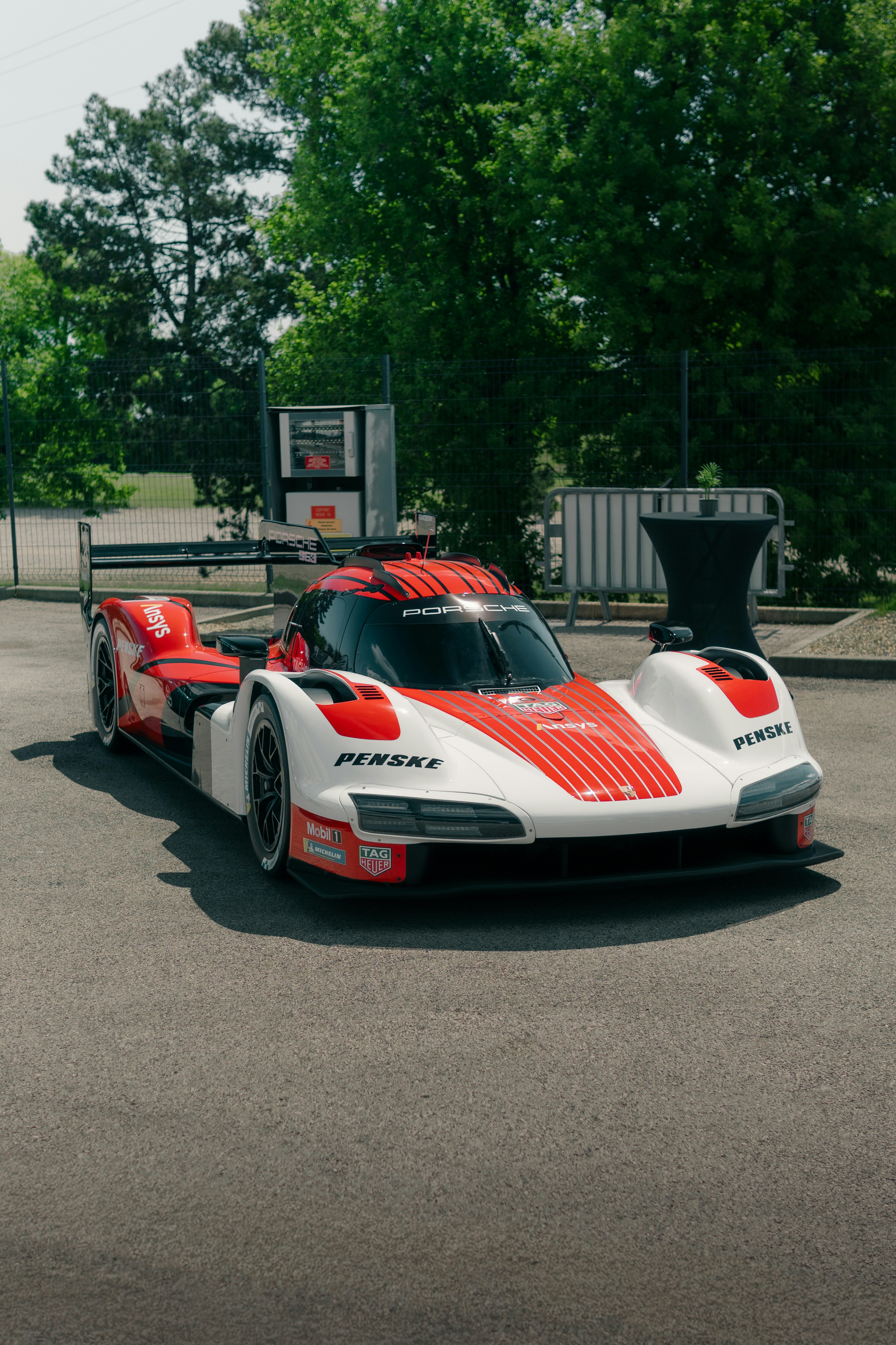 A sleek racing car with a striking red and white design, prominently featuring the Penske logo, parked against a backdrop of greenery.