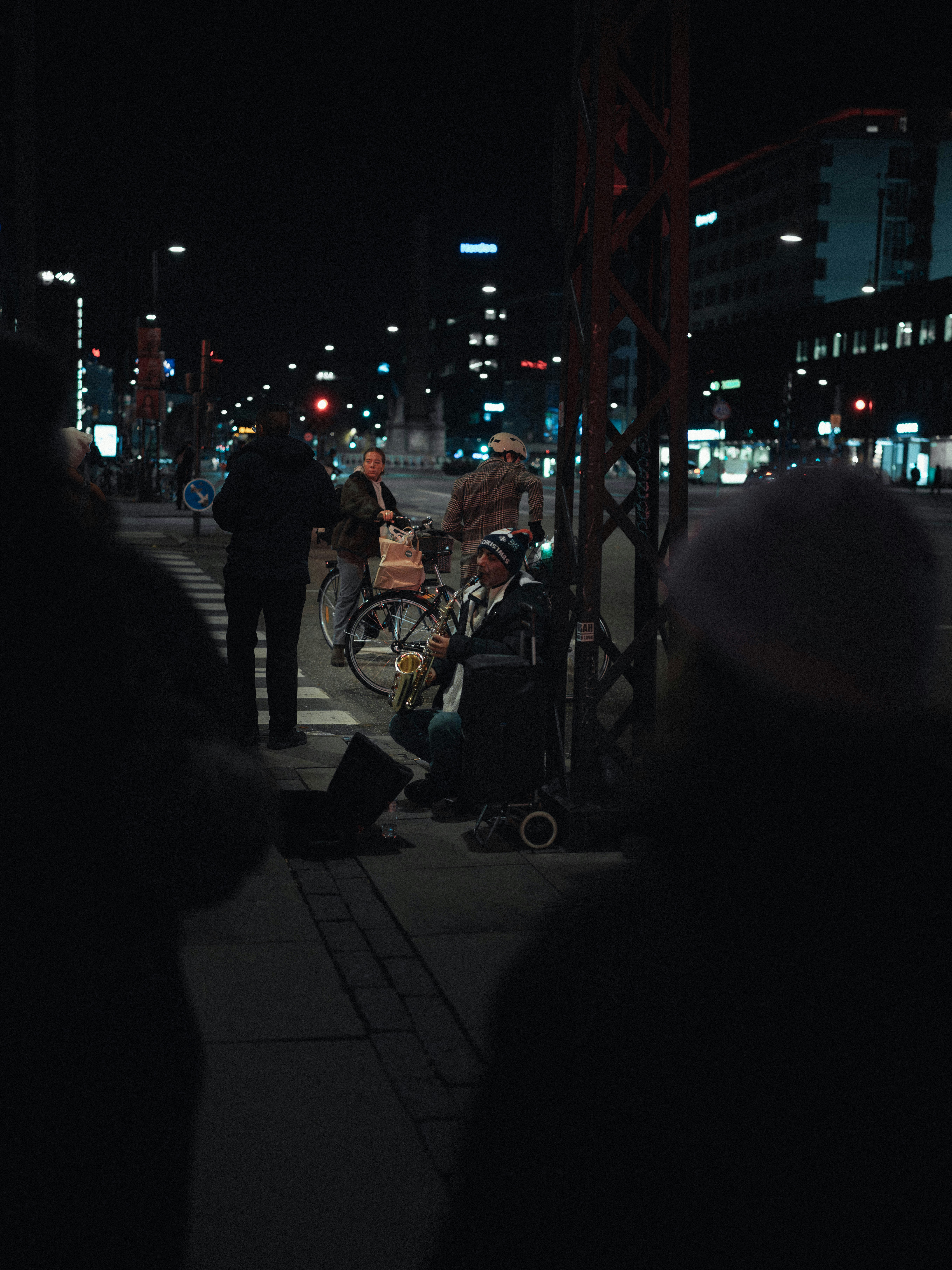 A street musician plays saxophone while a small group of people engages nearby, illuminated by urban lights at night.