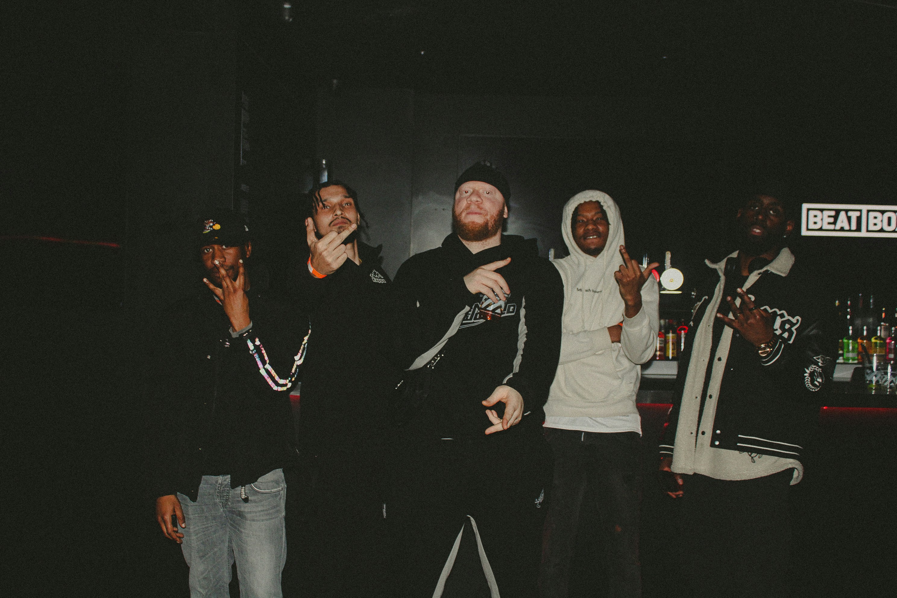 Five friends posing together in a dimly lit venue, showcasing their unique styles and camaraderie. The backdrop features a bar with illuminated bottles.