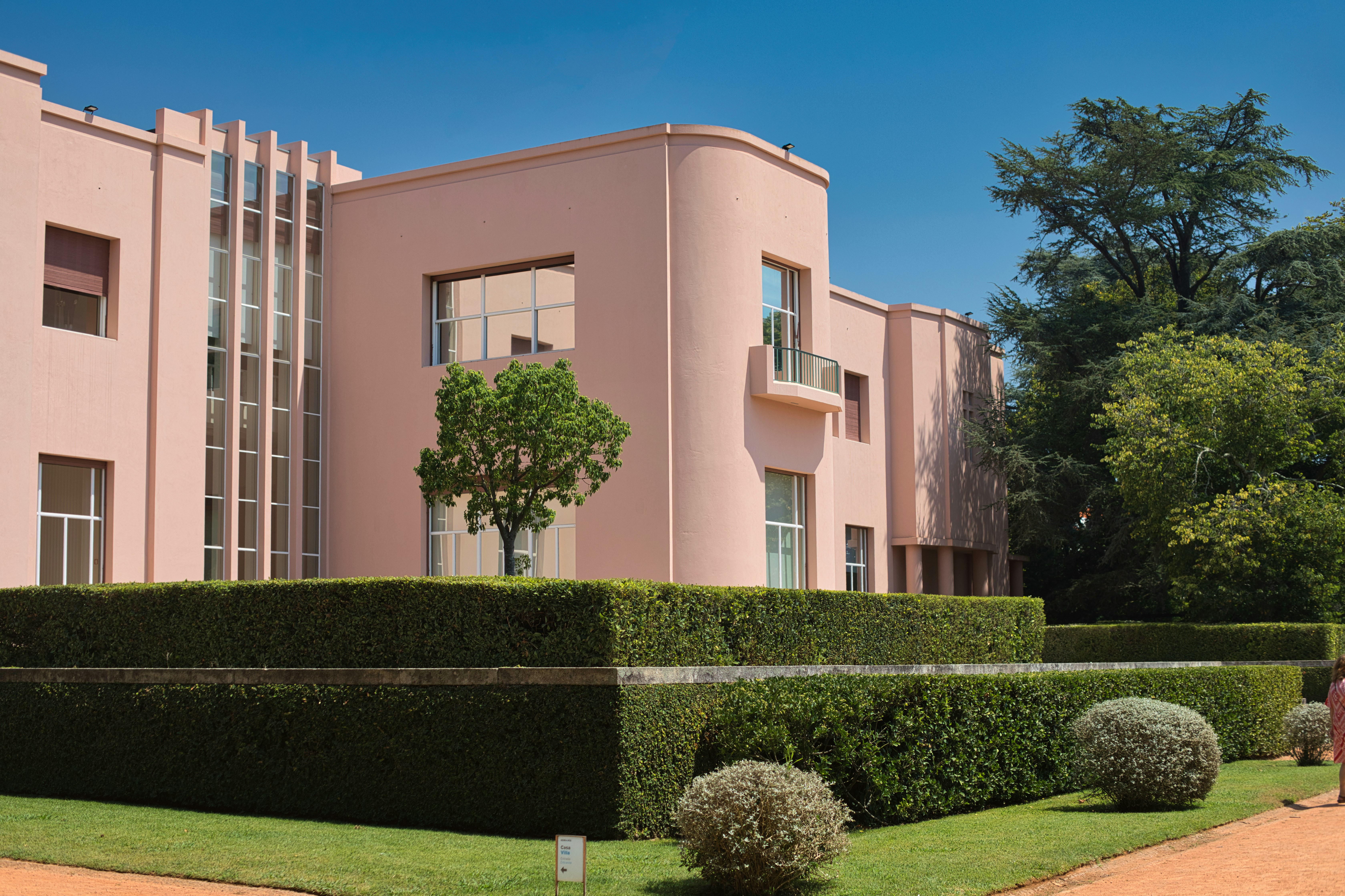 Modern architectural structure with pink walls and geometric design, surrounded by neatly trimmed hedges and greenery.