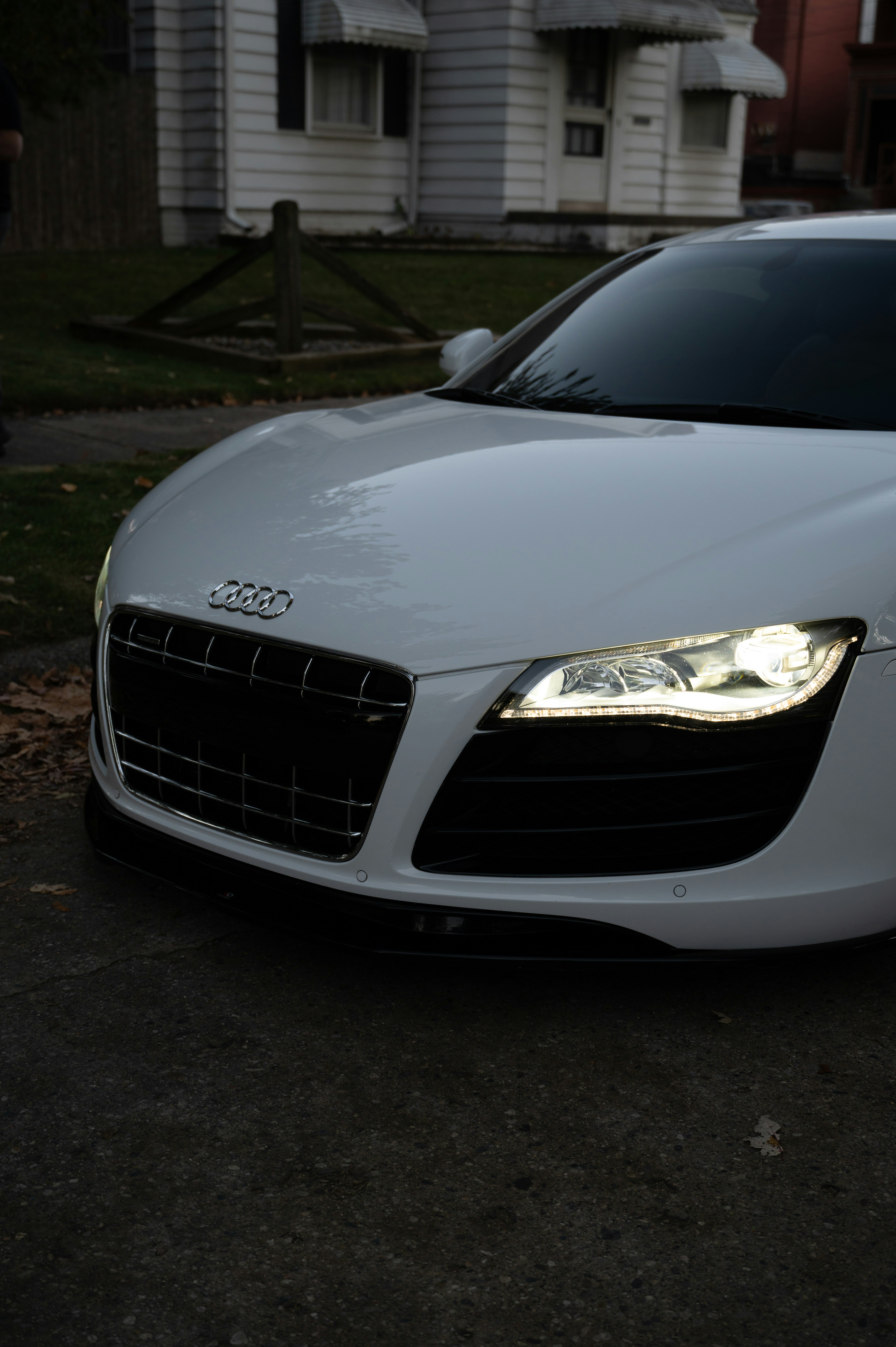 Sleek white Audi R8 parked on a quiet street, illuminated headlights casting a soft glow. The elegant design contrasts with the evening shadows.