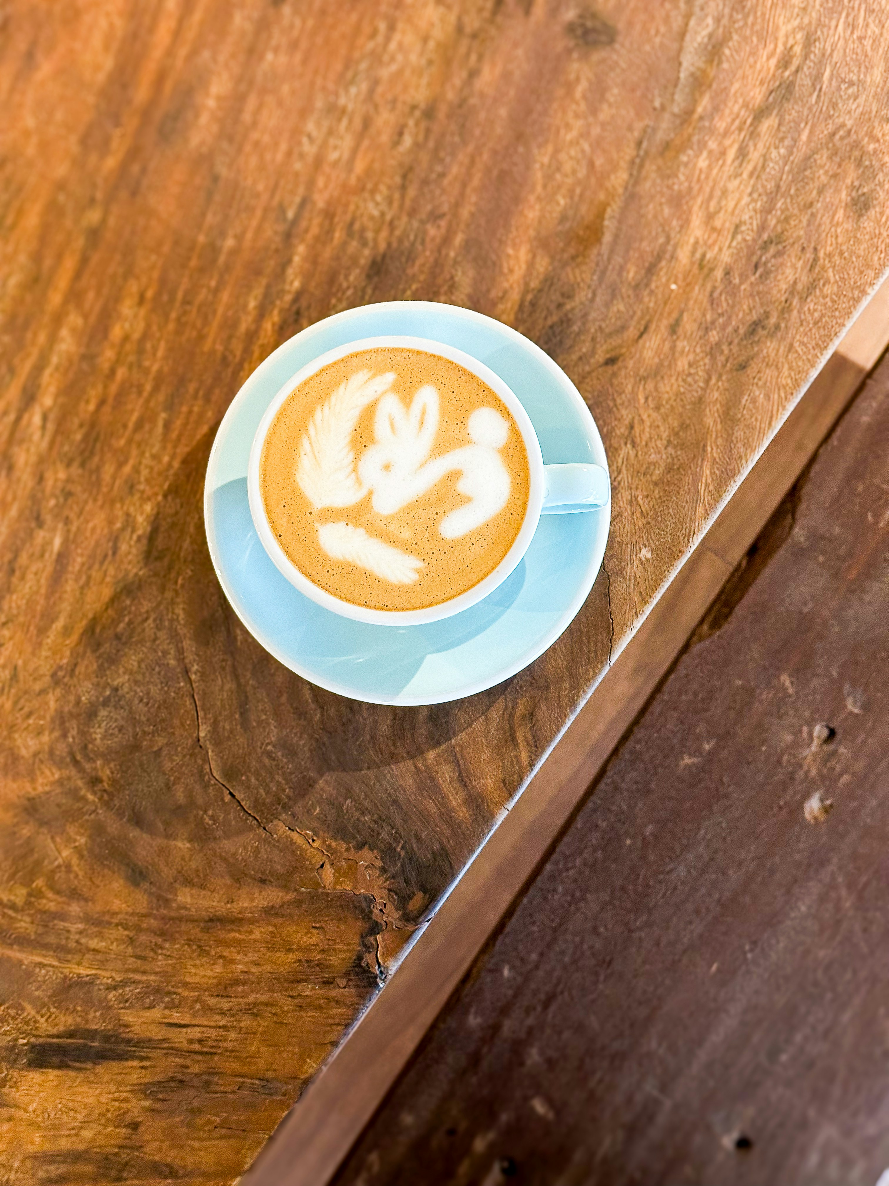 Latte art featuring a whimsical design of a rabbit and a person, served in a light blue cup on a wooden table.
