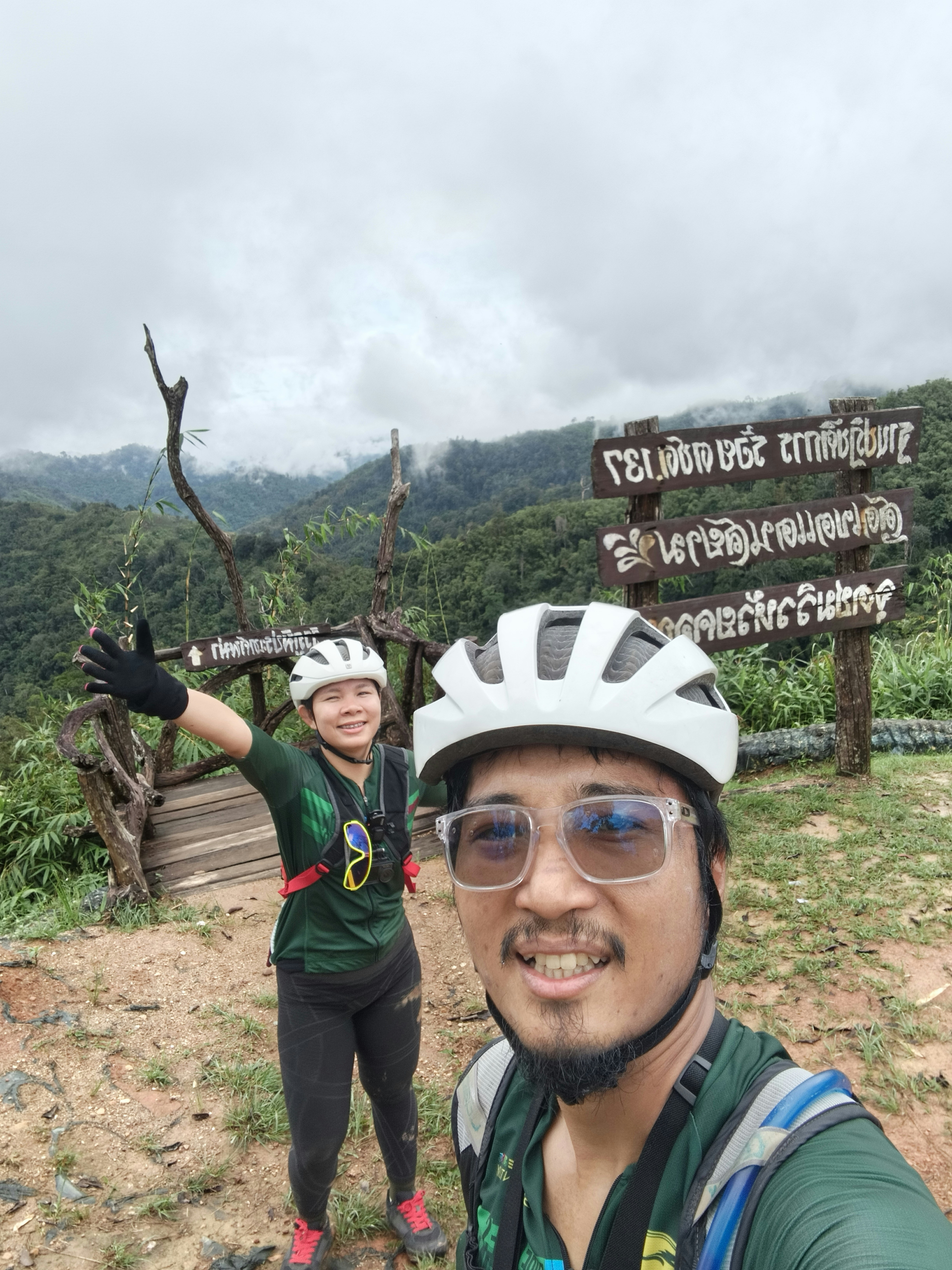 Ridge Day Selfie: Helmeted Duo Amid Carved Signposts on a Misty Mountain Trail