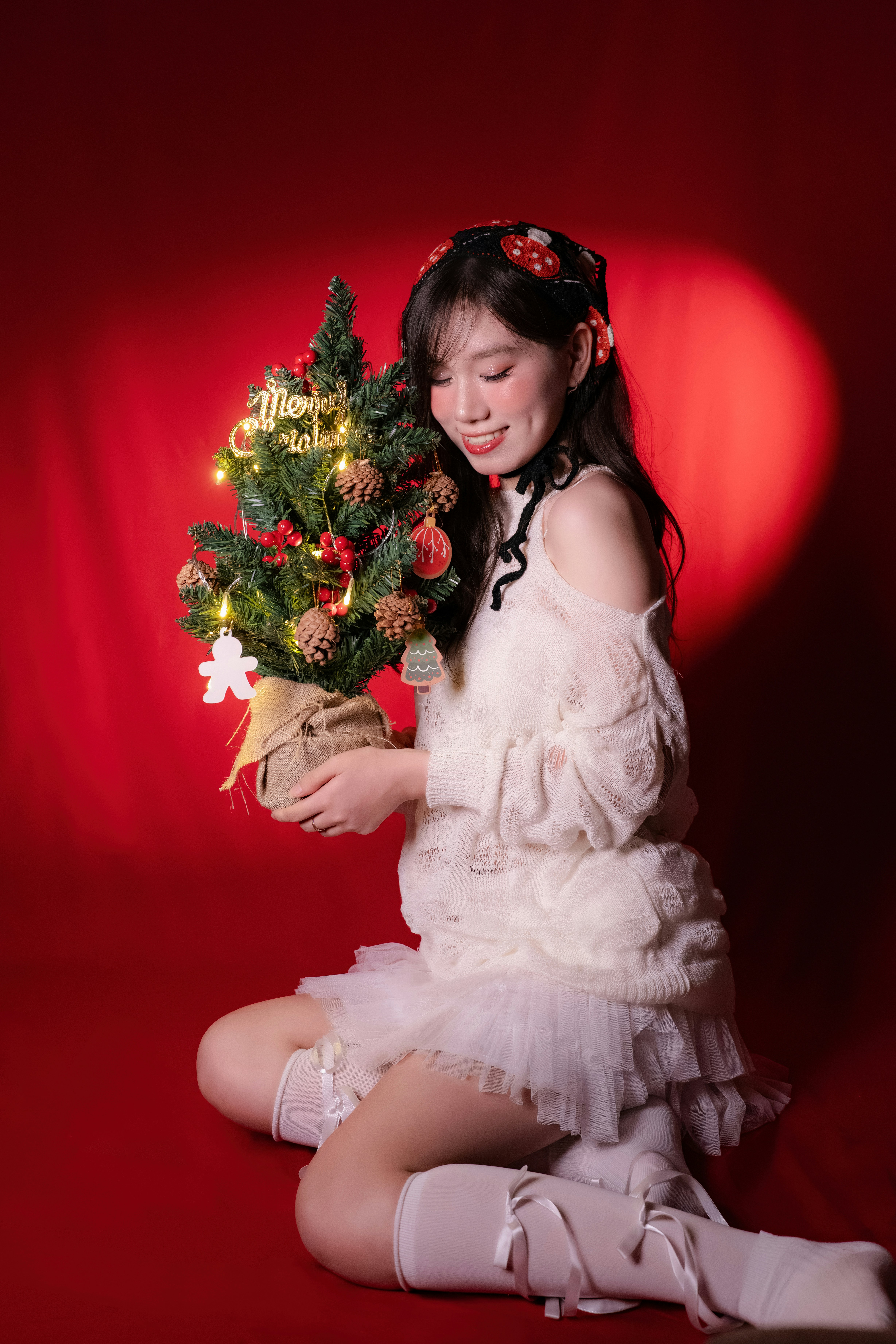Young woman in a cozy sweater holds a decorated mini Christmas tree against a vibrant red backdrop, radiating festive cheer.