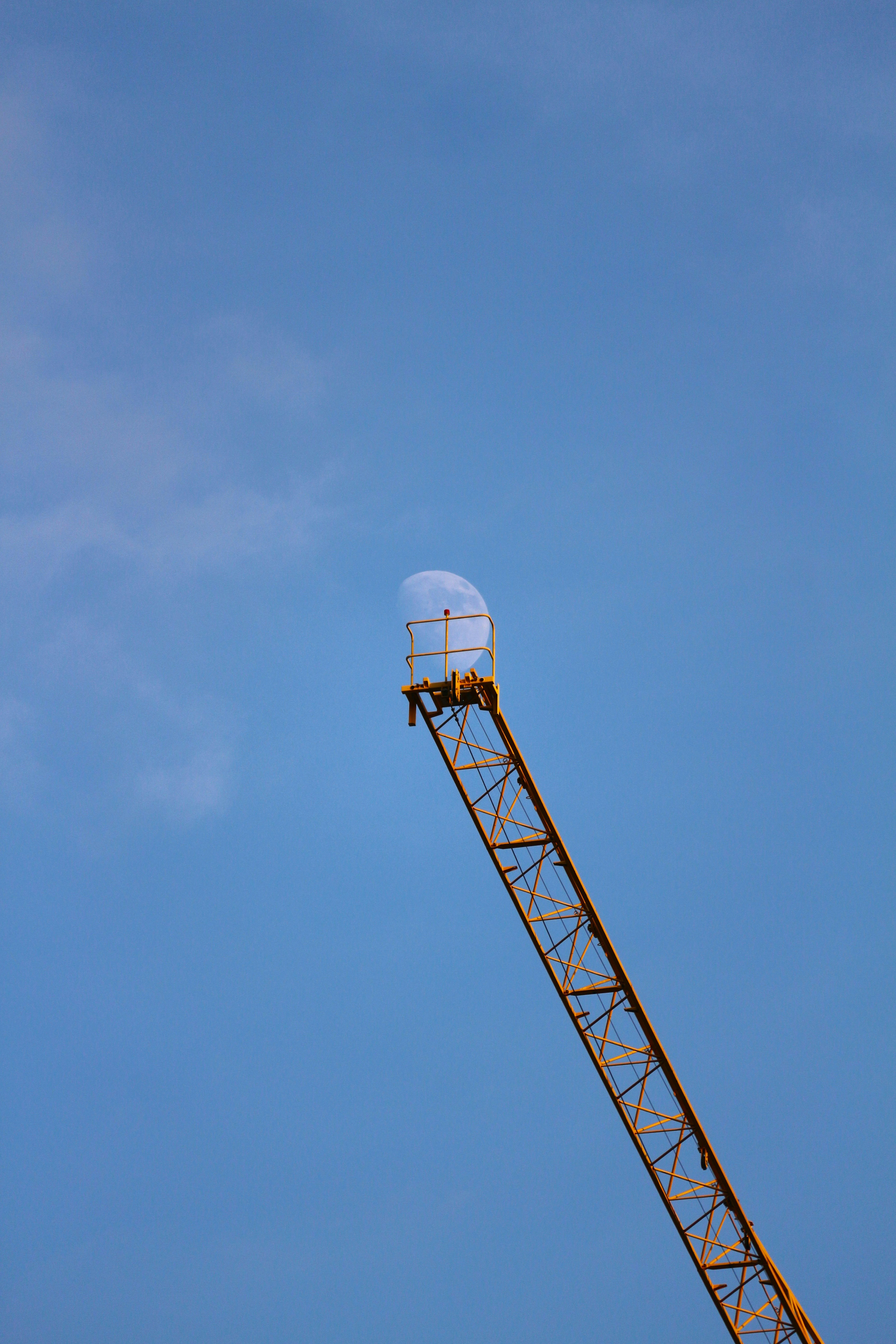 A towering crane extends toward the sky, its silhouette contrasted against a blue backdrop with wispy clouds. The moon peeks over the top, adding a whimsical touch.