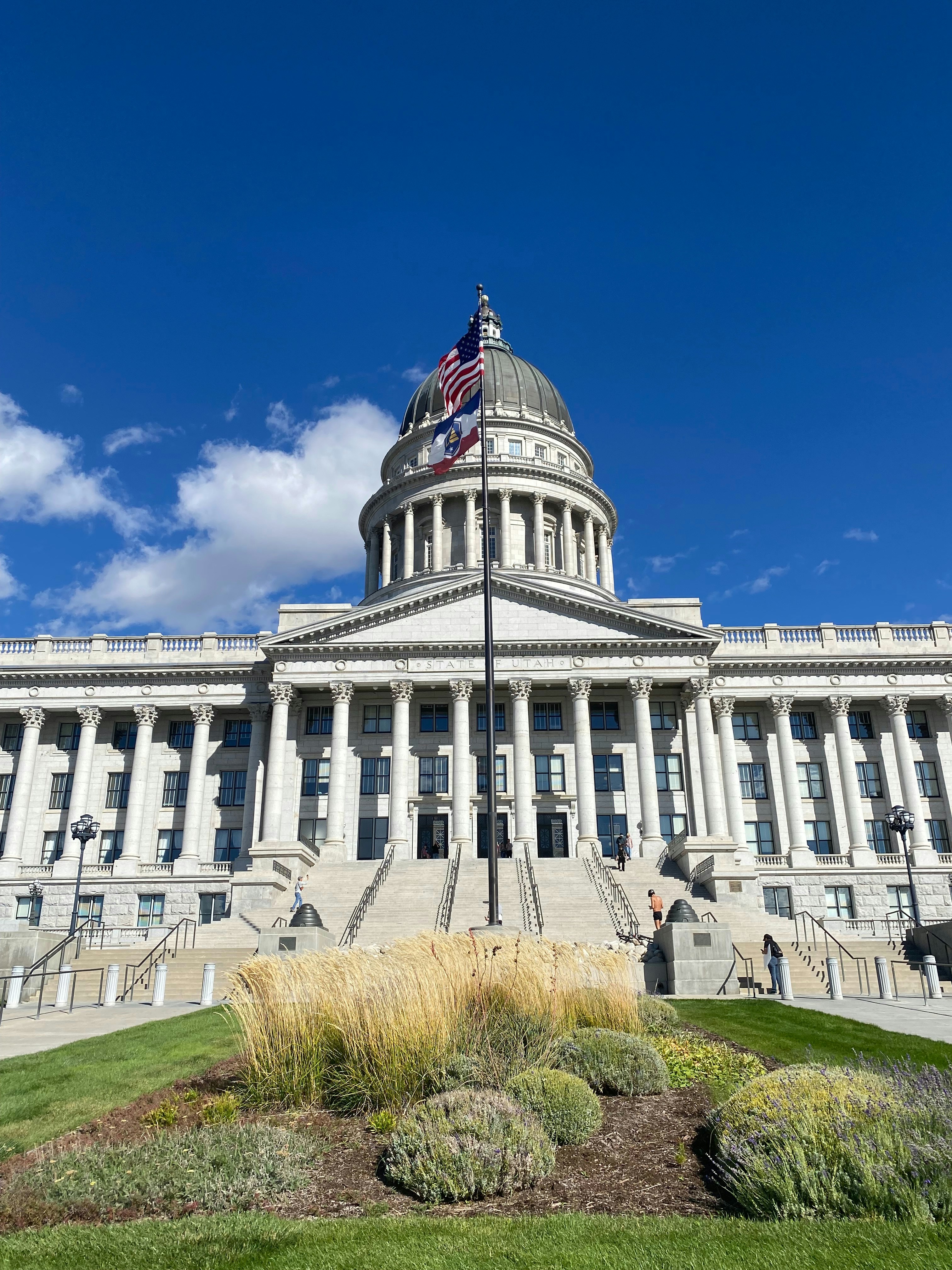 The grand steps of a government building lead up to an impressive dome, with flags waving gently in the breeze against a vivid blue sky.