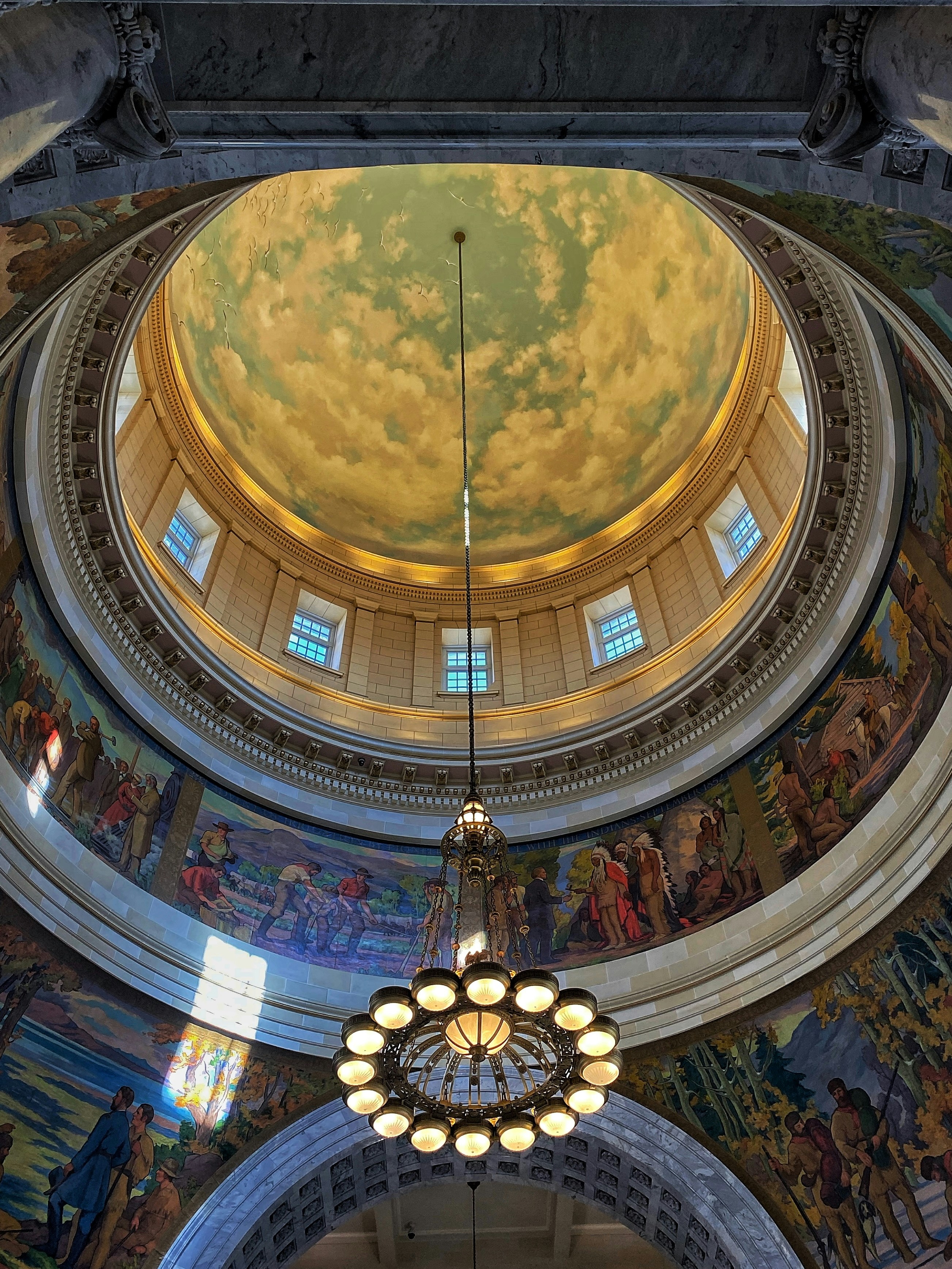 Intricate murals and a luminous chandelier adorn the grand dome of a historic building, showcasing artistic mastery and architectural elegance.