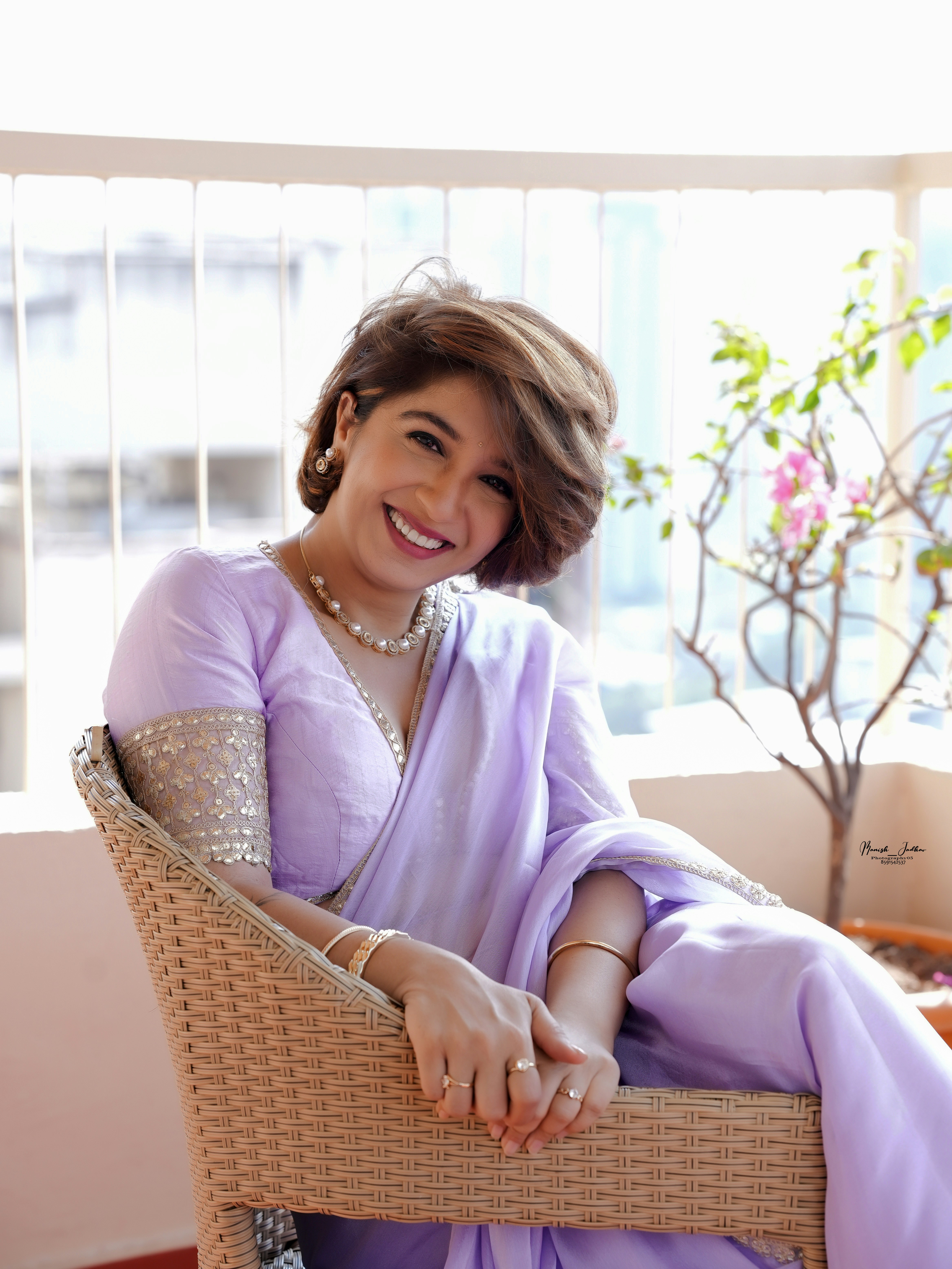 A woman in a lavender sari smiles warmly while seated in a wicker chair, with a flowering plant in the background. The soft lighting enhances her joyful expression.