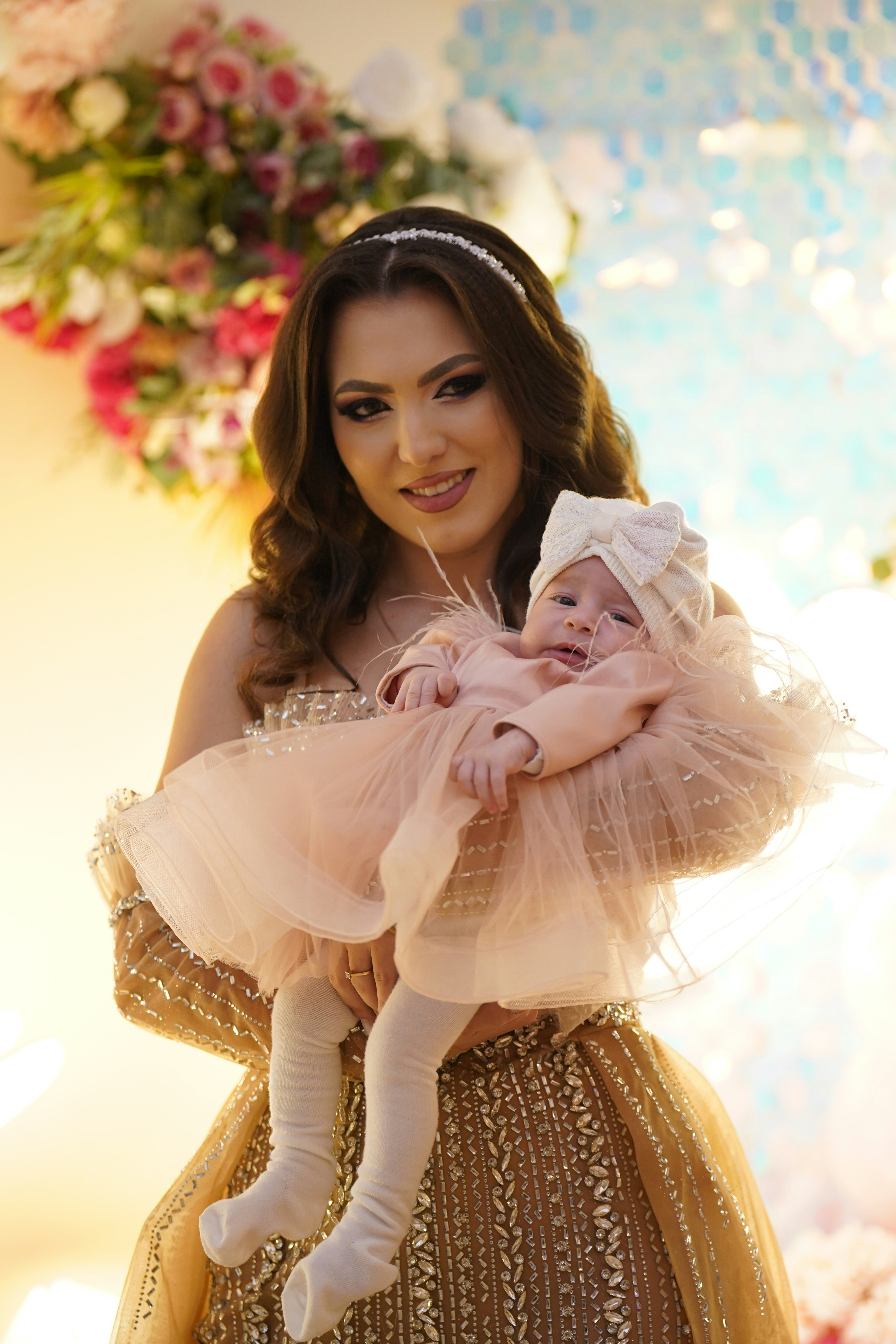 Woman in a shimmering gown cradling an infant dressed in a delicate outfit, surrounded by floral decorations.