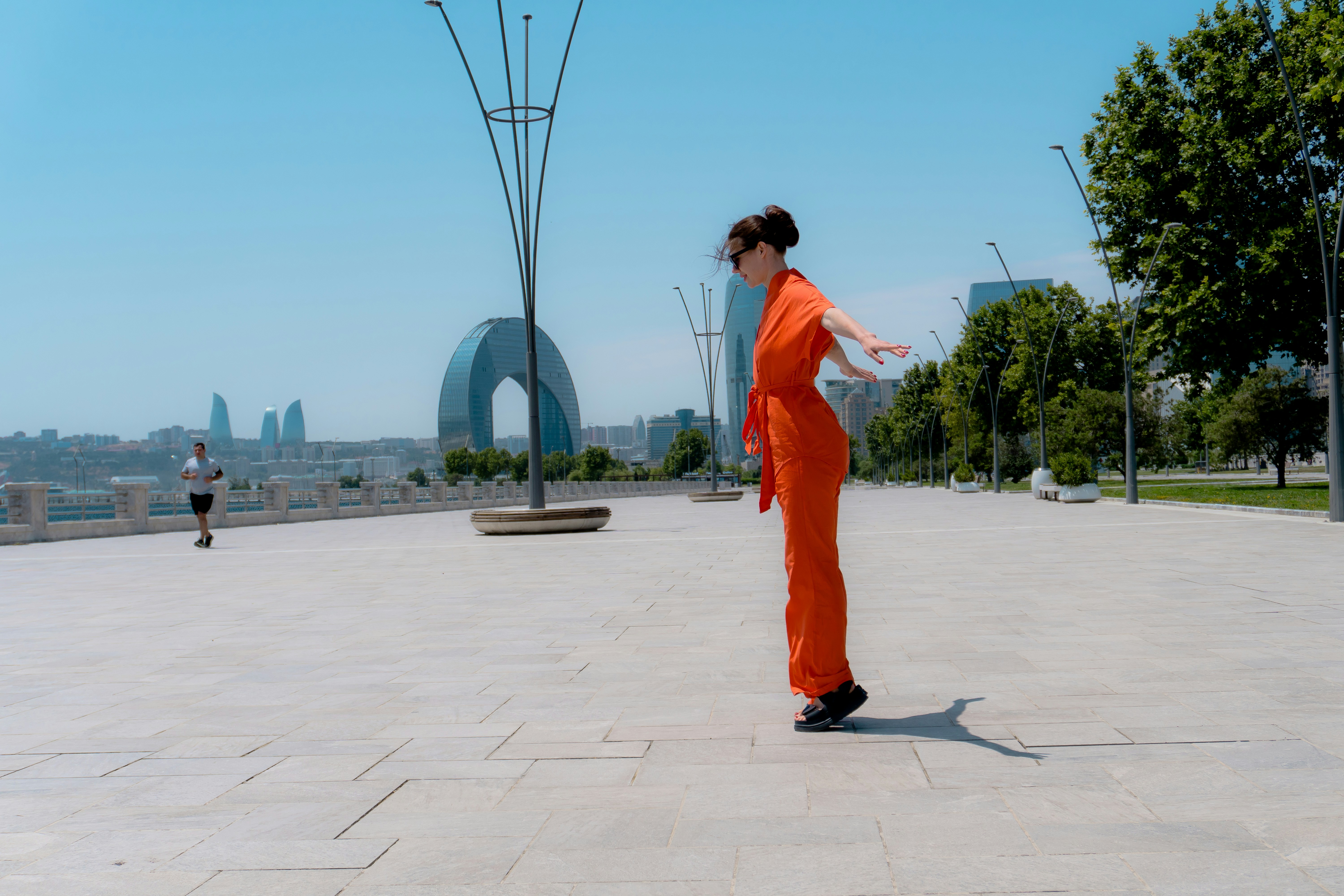 A woman in a vibrant orange outfit stands poised on a city promenade, with modern architecture and greenery in the background.
