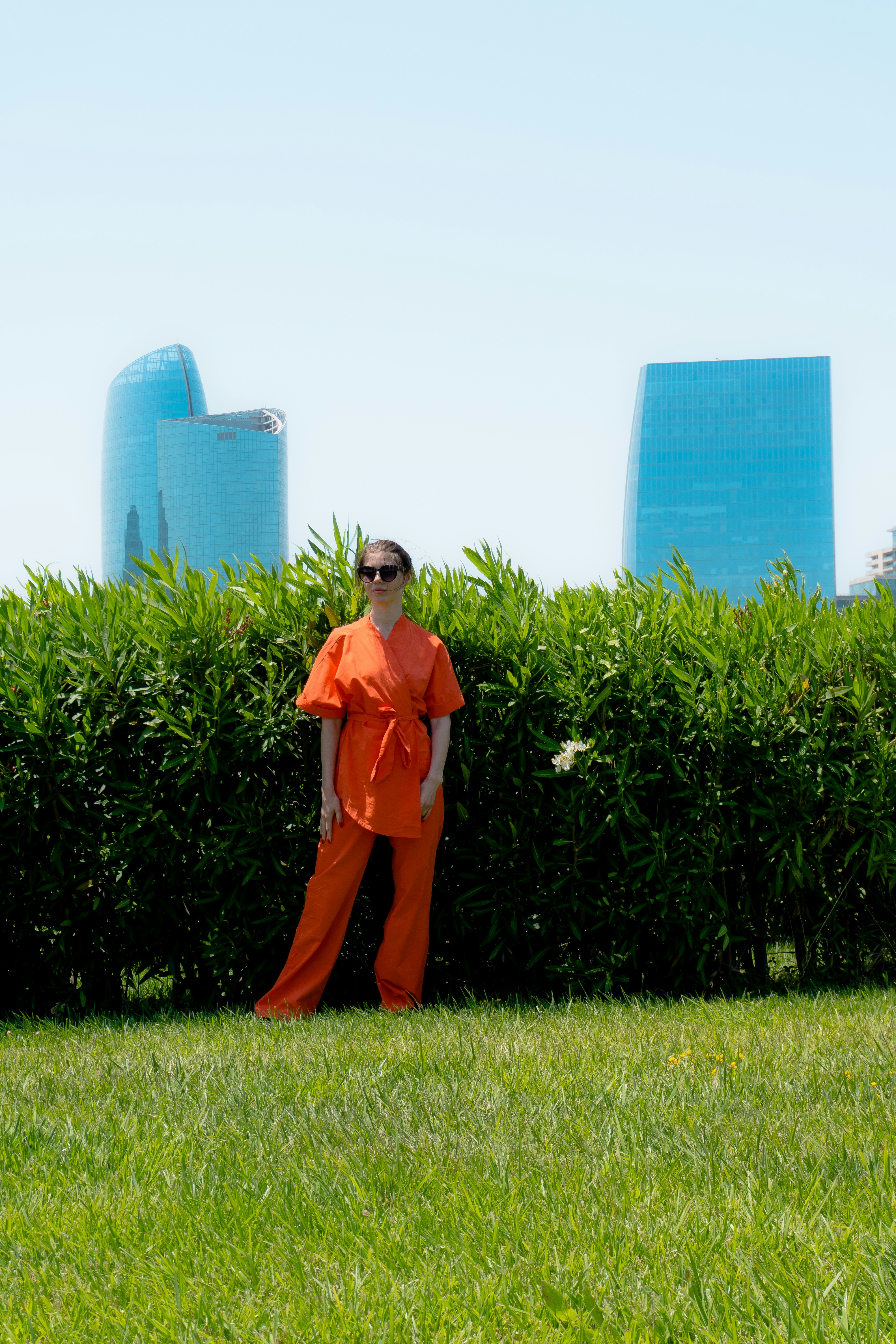 Orange-clad figure anchors a hedge wall against blue-sky skyscrapers