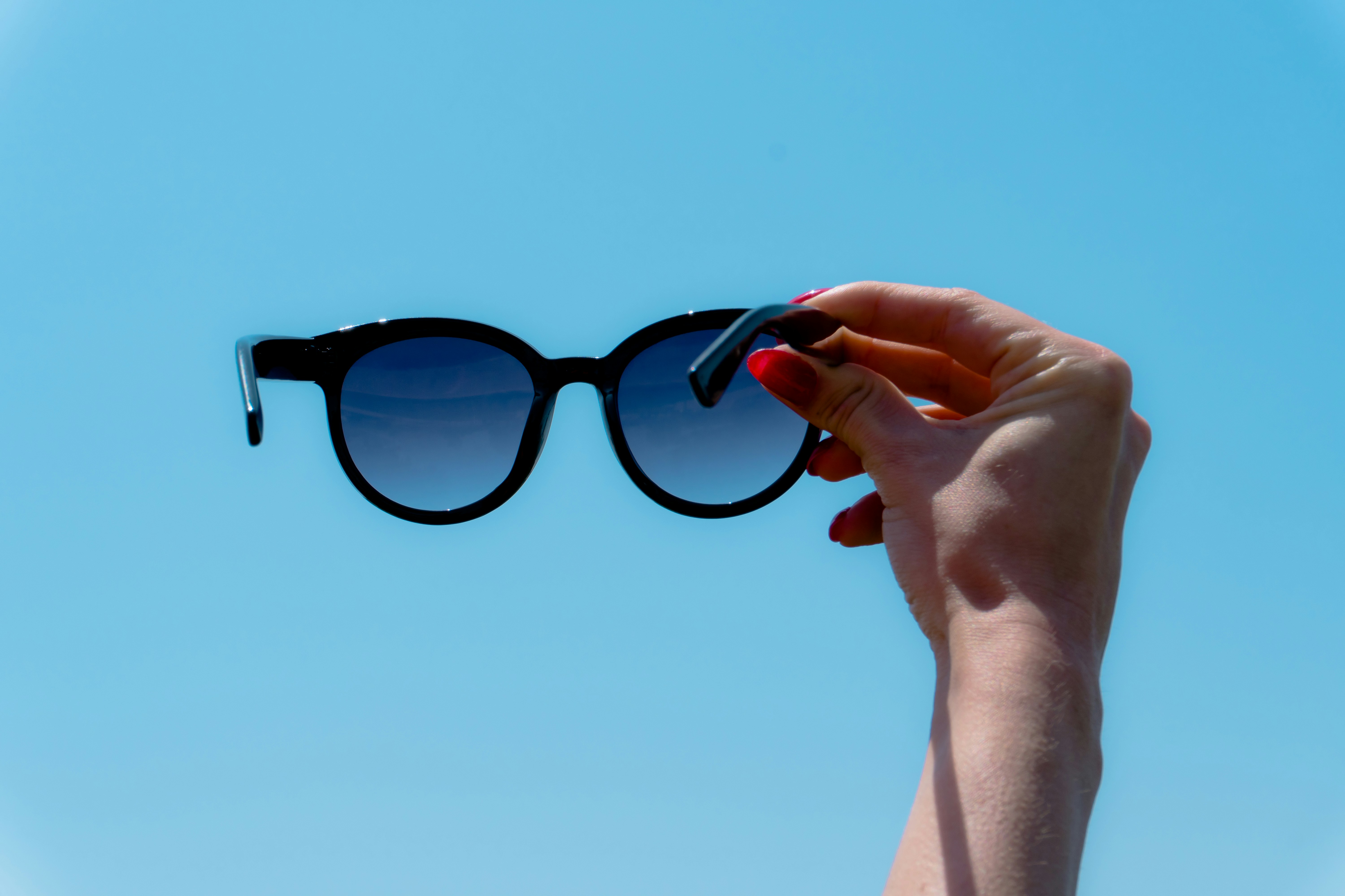 Black sunglasses held against a clear blue sky, highlighting the contrast between the accessory and the vibrant backdrop.
