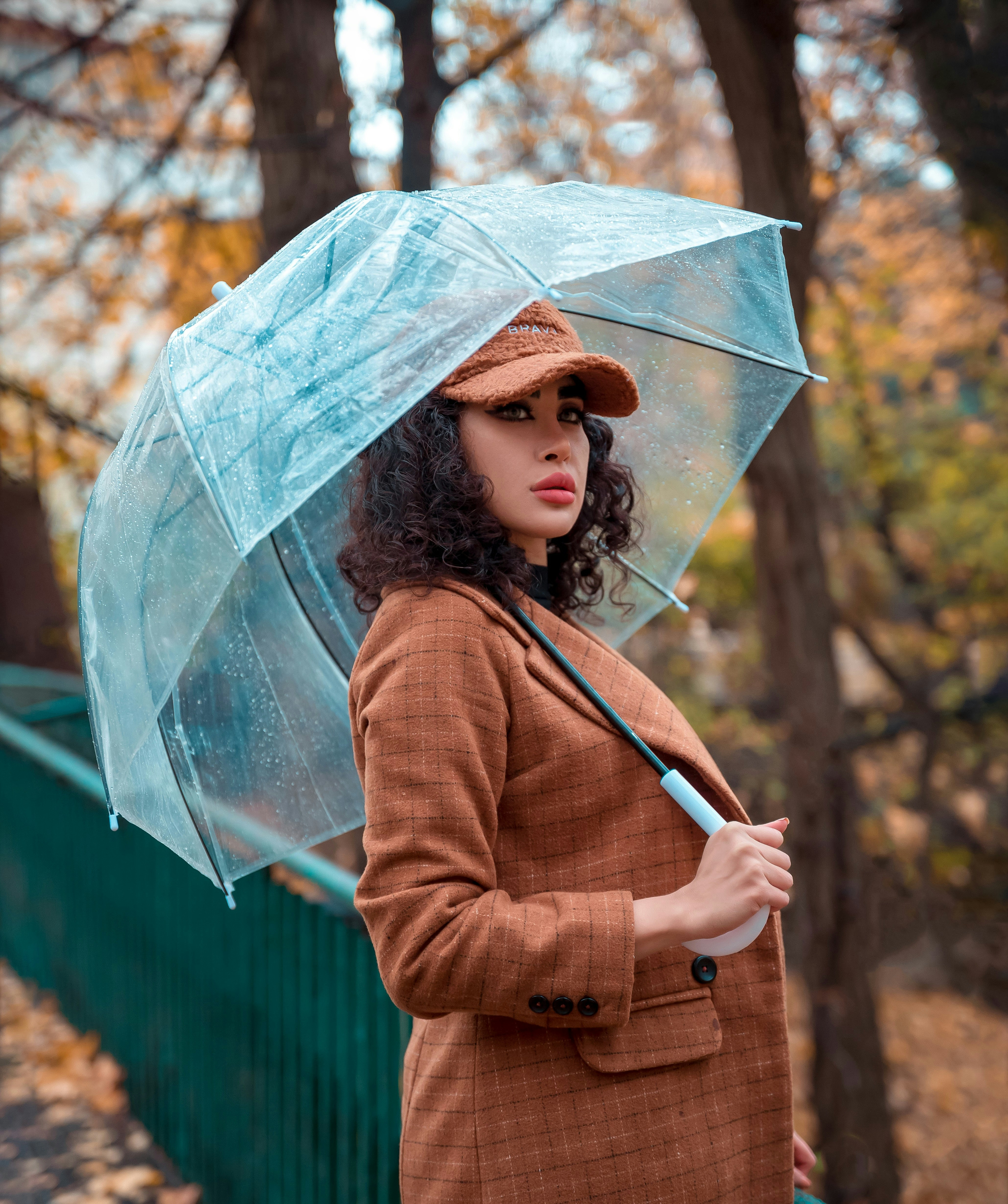 A woman in a stylish brown coat holds a clear umbrella, surrounded by autumn foliage. The scene captures a serene moment amidst light rain.
