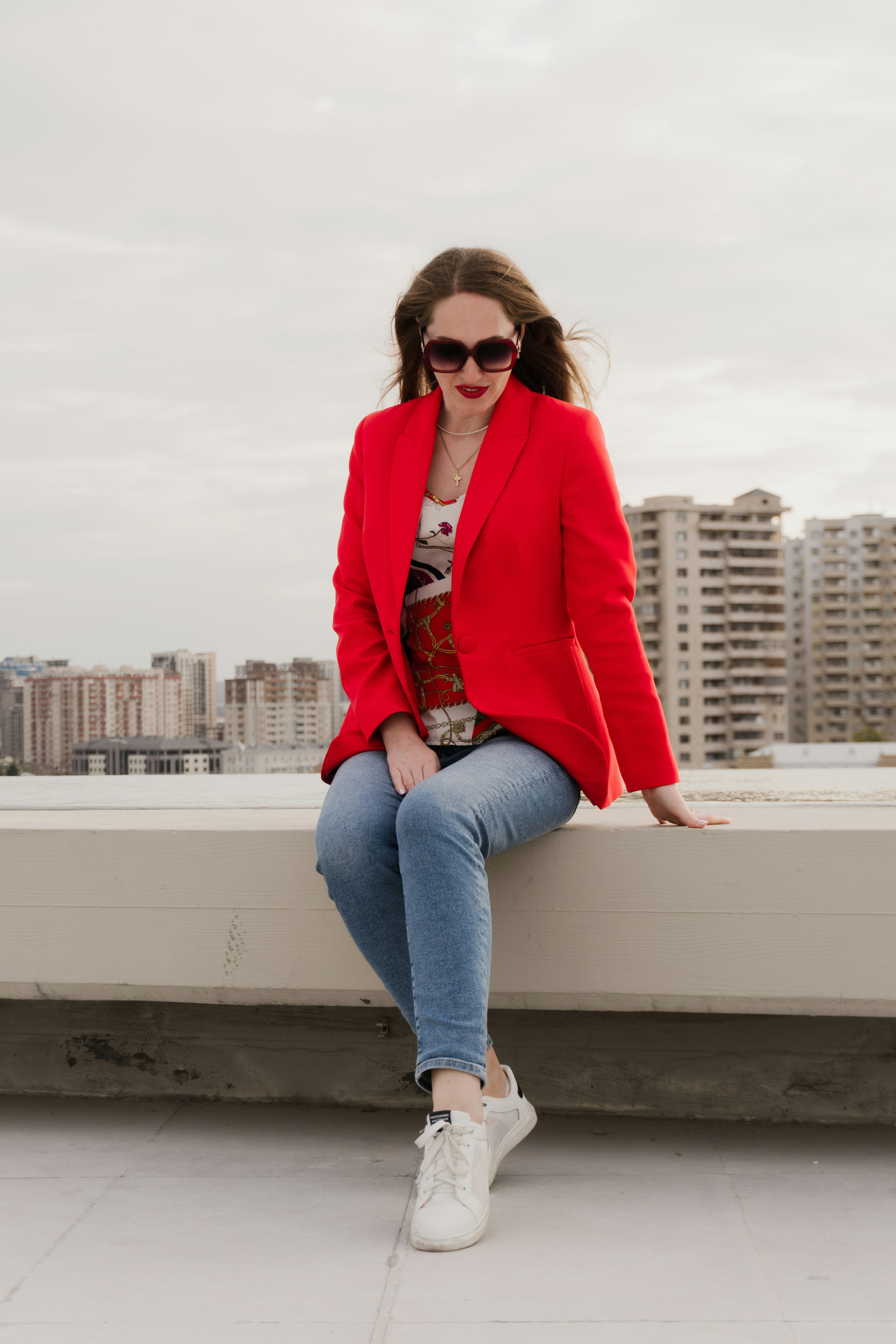 Woman in a vibrant red blazer and sunglasses sits on a rooftop ledge, overlooking a cityscape of modern buildings. Wind tousles her hair.