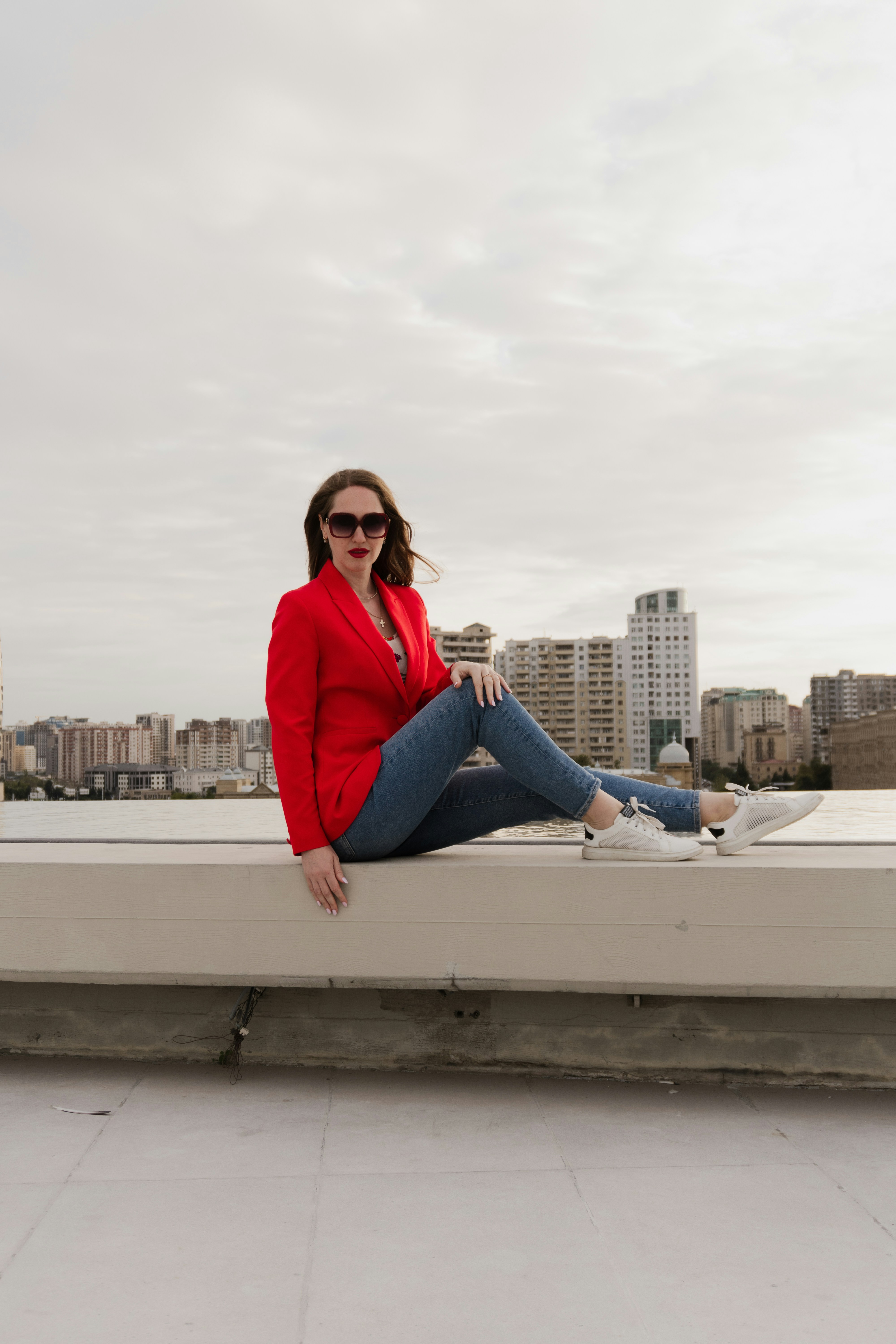 Woman in a red blazer and sunglasses poses on a rooftop against a city skyline backdrop.