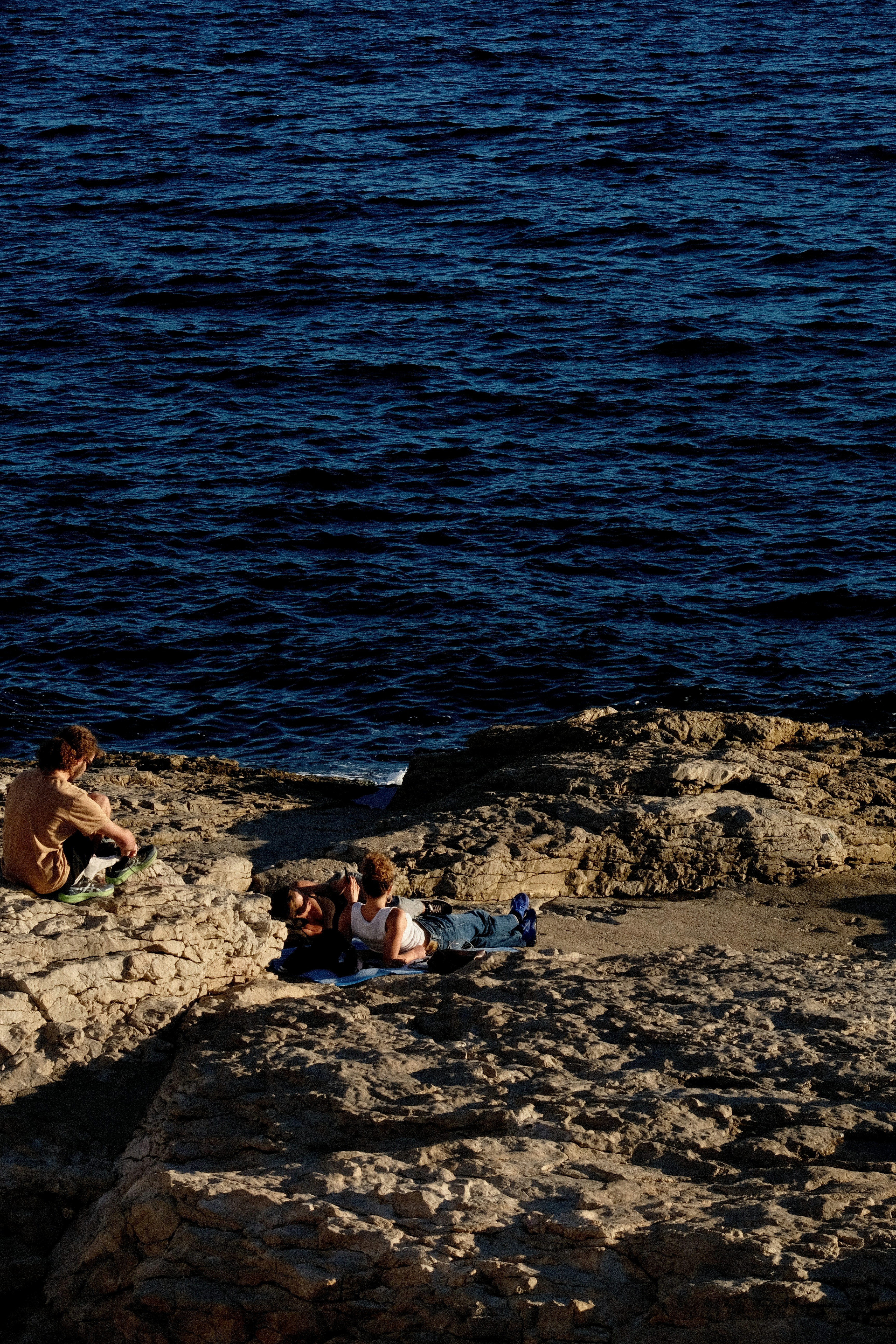 Two people relaxing on rocky shore by dark blue ocean. photo – Free ...