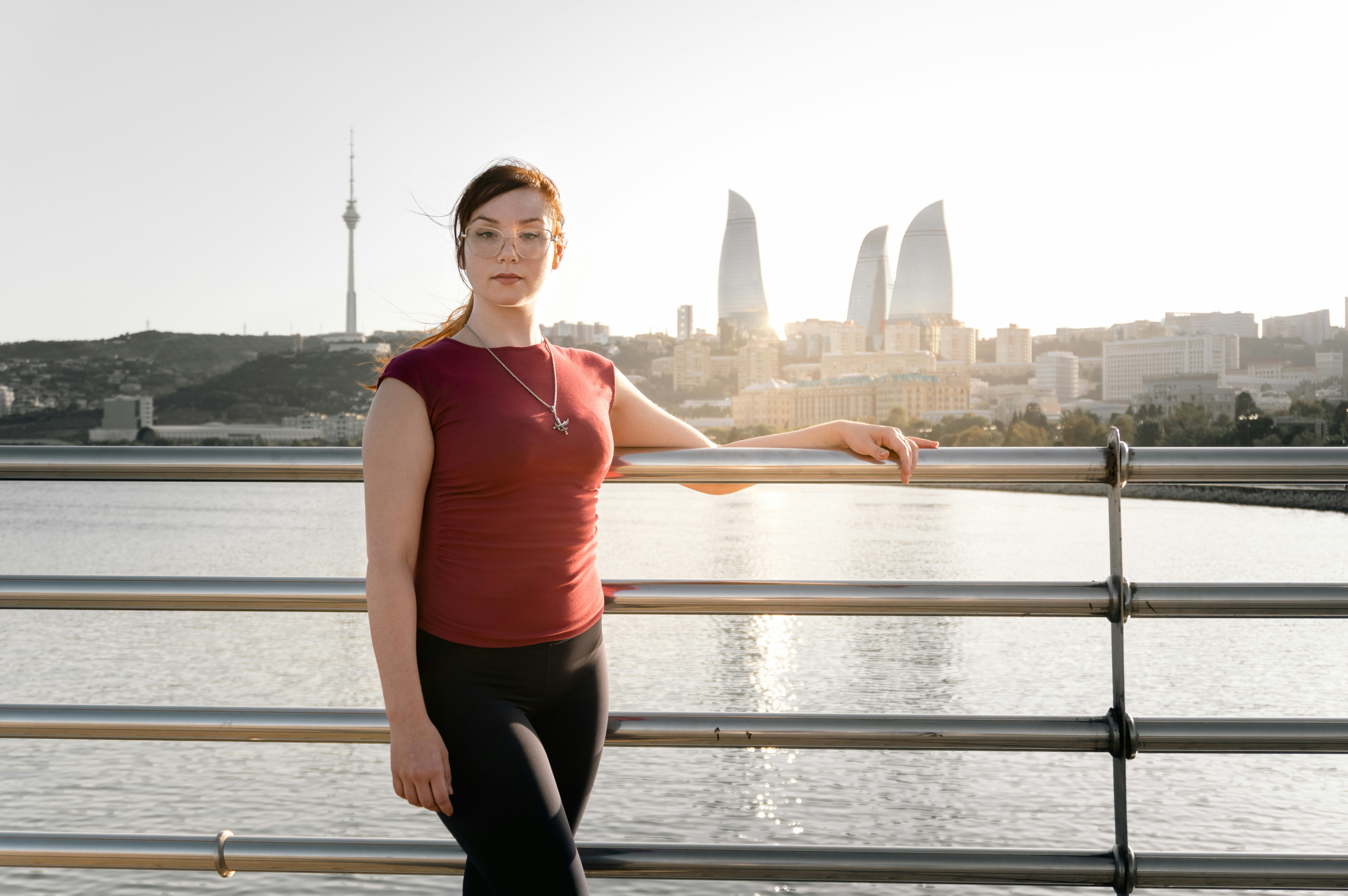 A woman in a fitted red top poses by a railing with a city skyline in the background, showcasing a blend of natural and urban elements.
