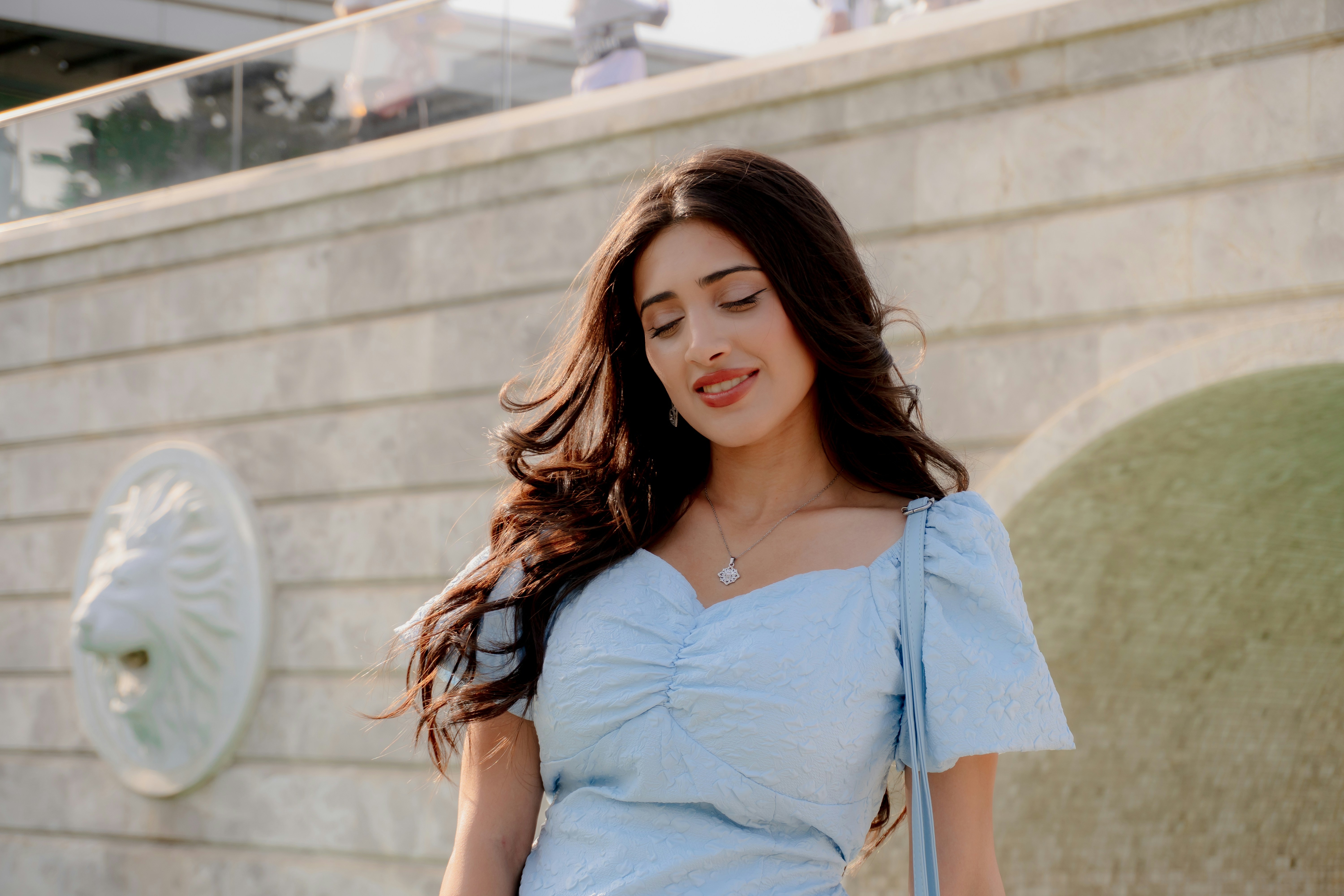A woman in a light blue dress poses gracefully near a decorative stone wall with a lion motif.