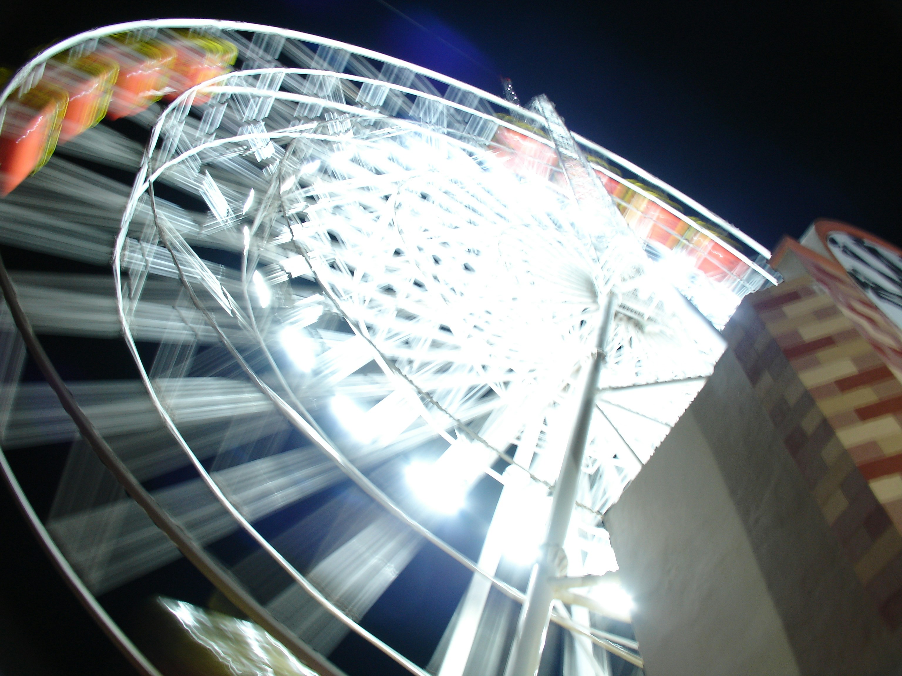 A vibrant Ferris wheel illuminated by bright lights spins against the dark sky, conveying the energy of a night fair.