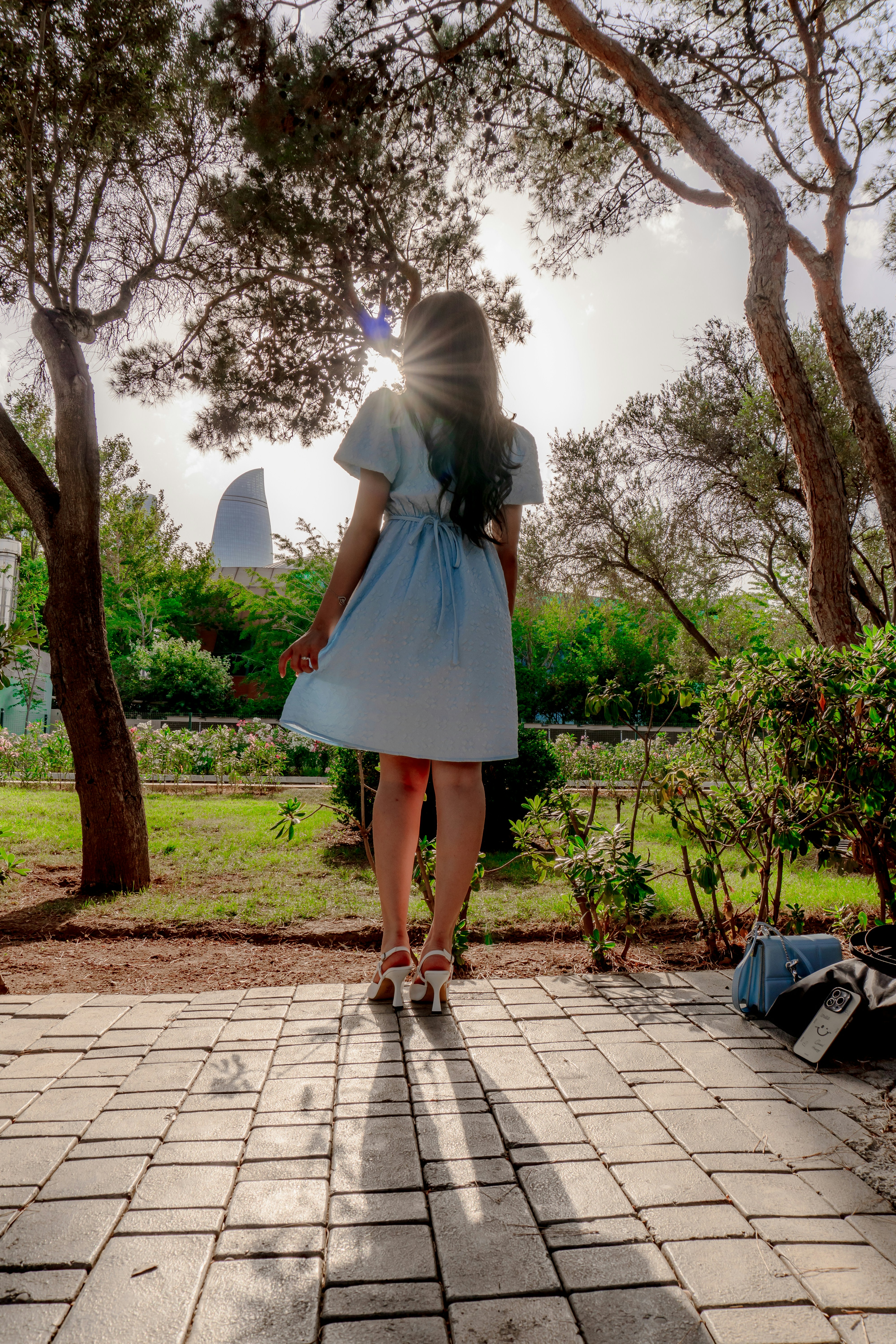 A woman in a light blue dress stands in a lush garden, sunlight filtering through the trees, casting intricate shadows on the ground.