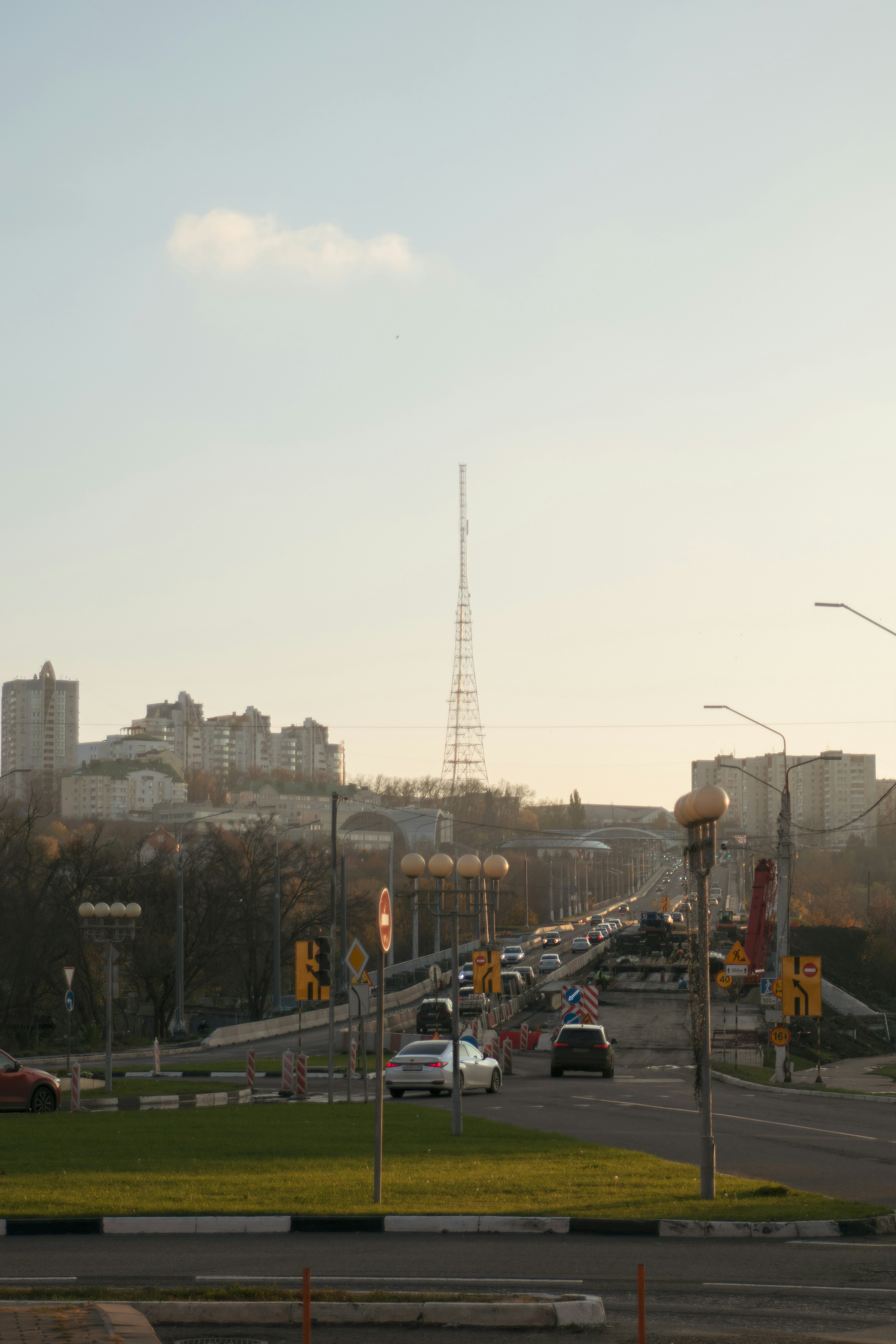 A busy urban road lined with traffic, leading towards a distant bridge and communication tower, framed by residential buildings. The scene captures the essence of city life.