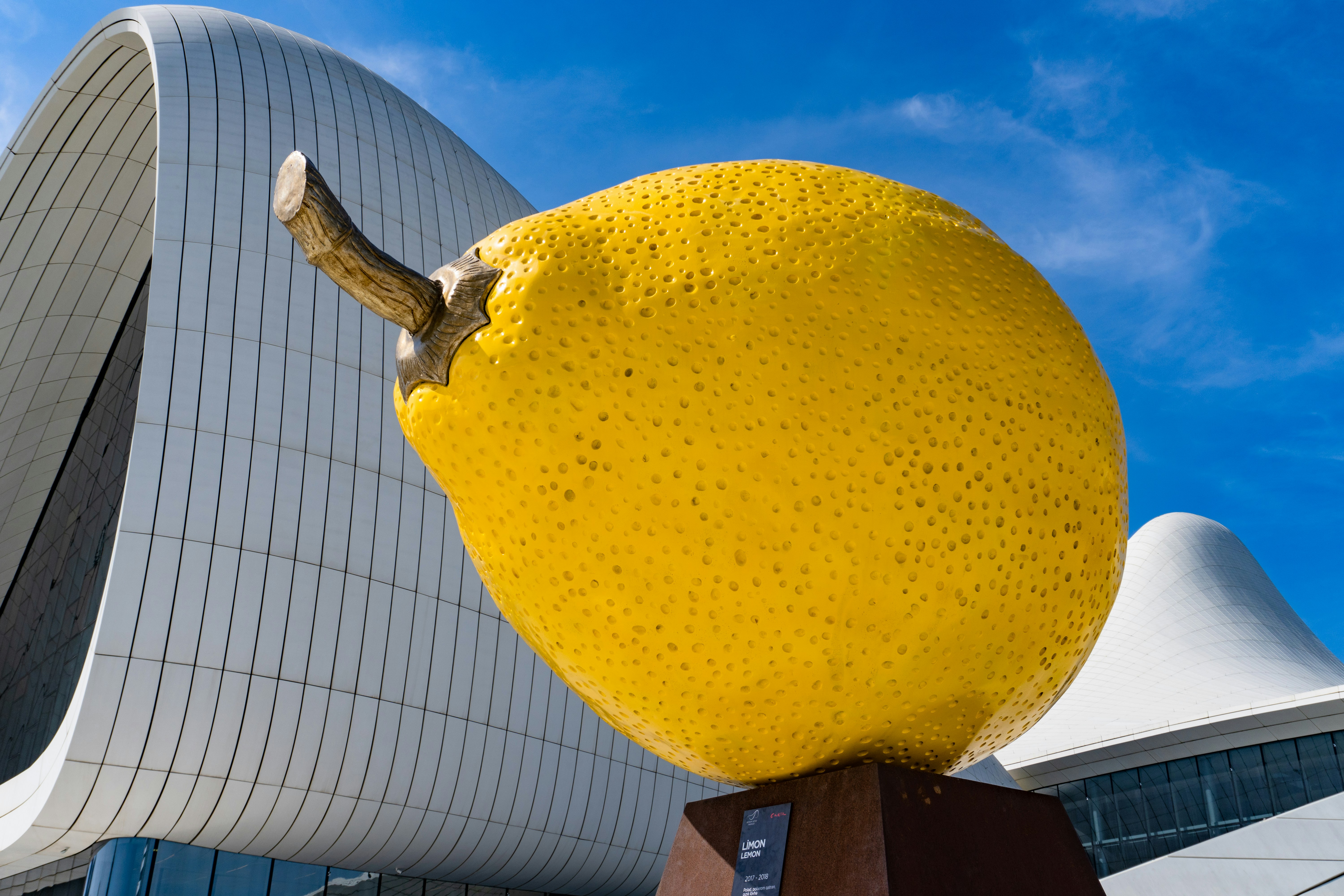 Oversized yellow lemon sculpture stands prominently in front of modern architectural structures under a bright blue sky.