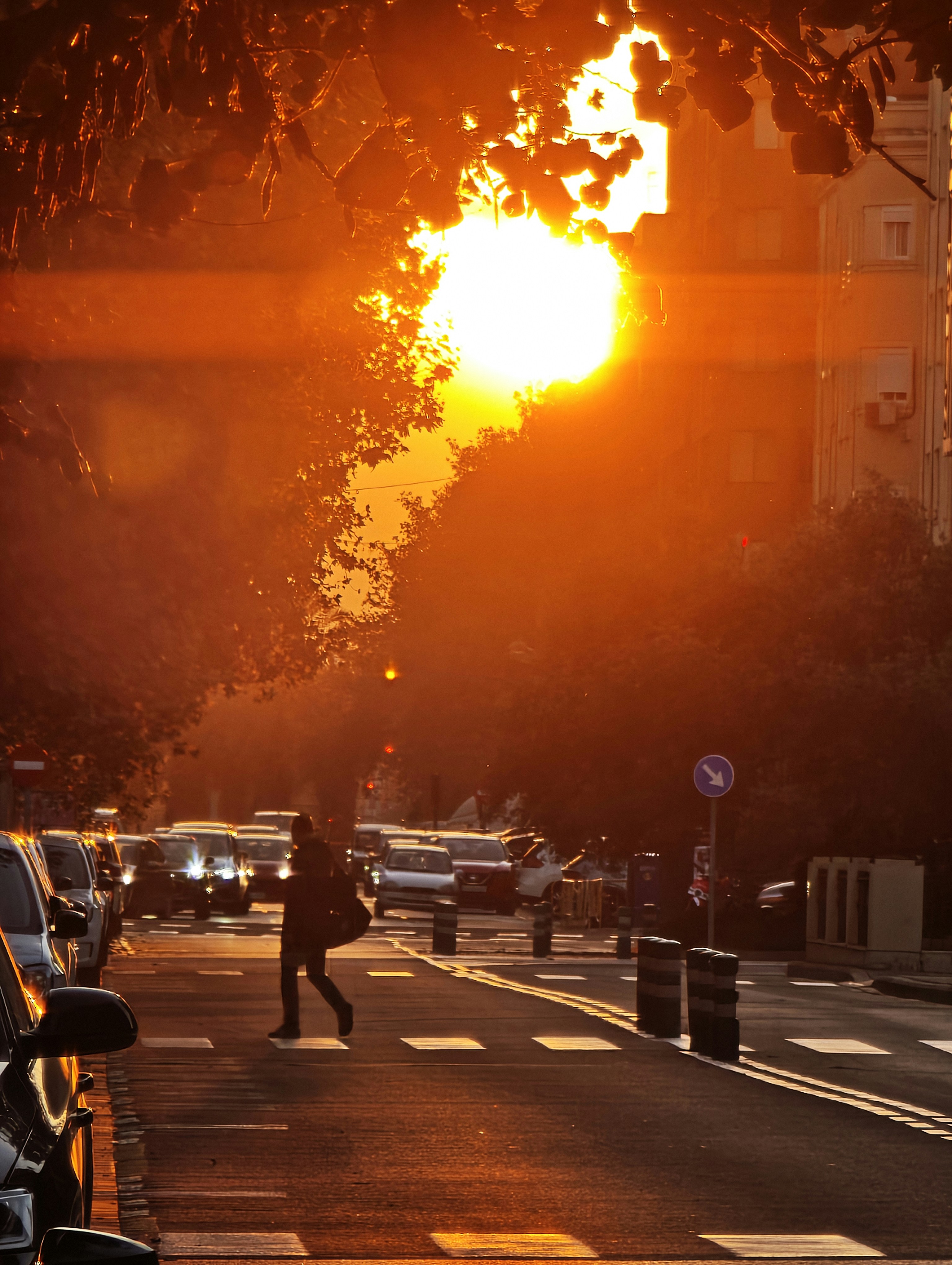 Silhouette of a pedestrian crossing a sunlit street, framed by vehicles and illuminated by a vibrant sunset.
