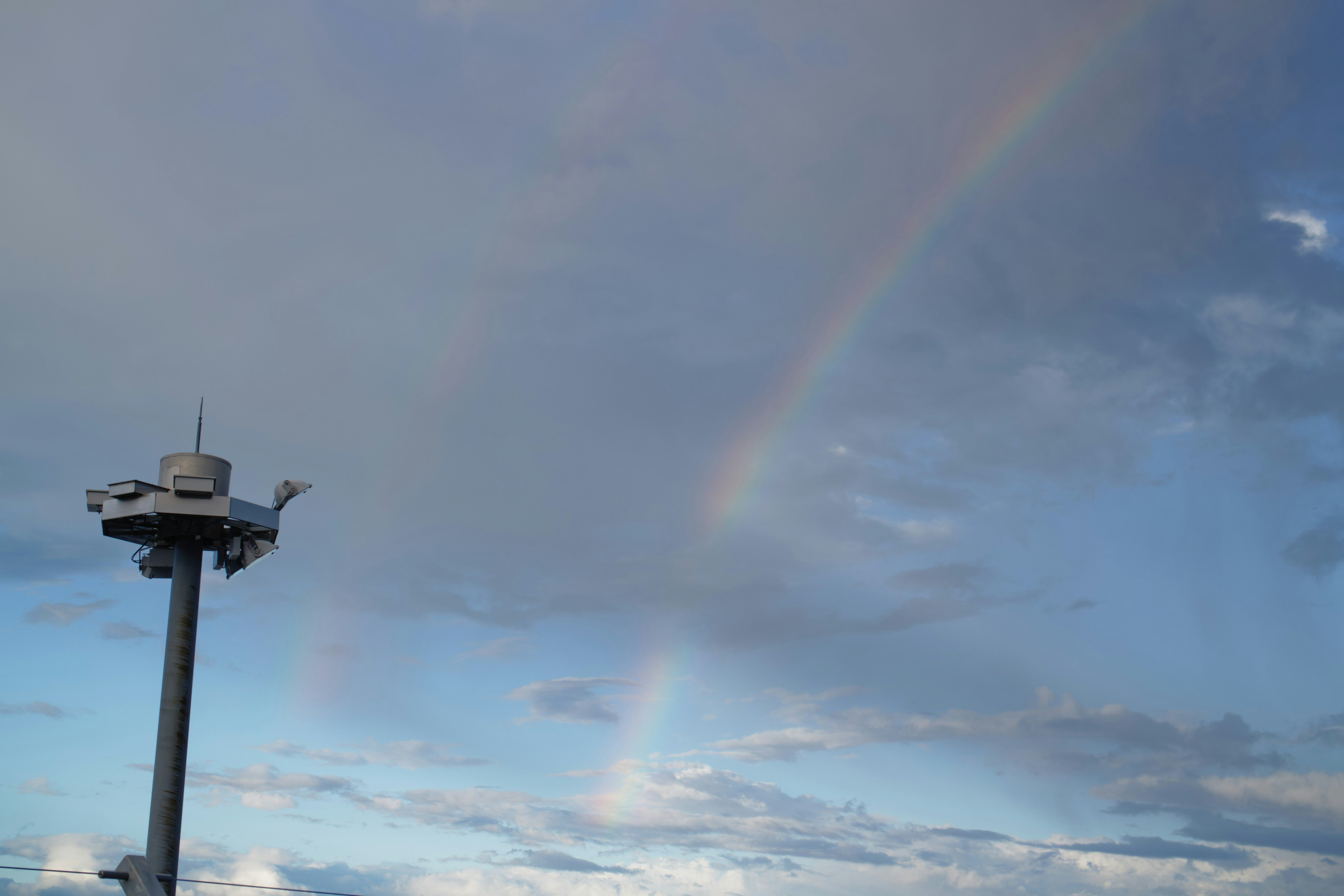 Double Rainbow Framing a Tall Surveillance Mast Under a Cloudy Blue Sky