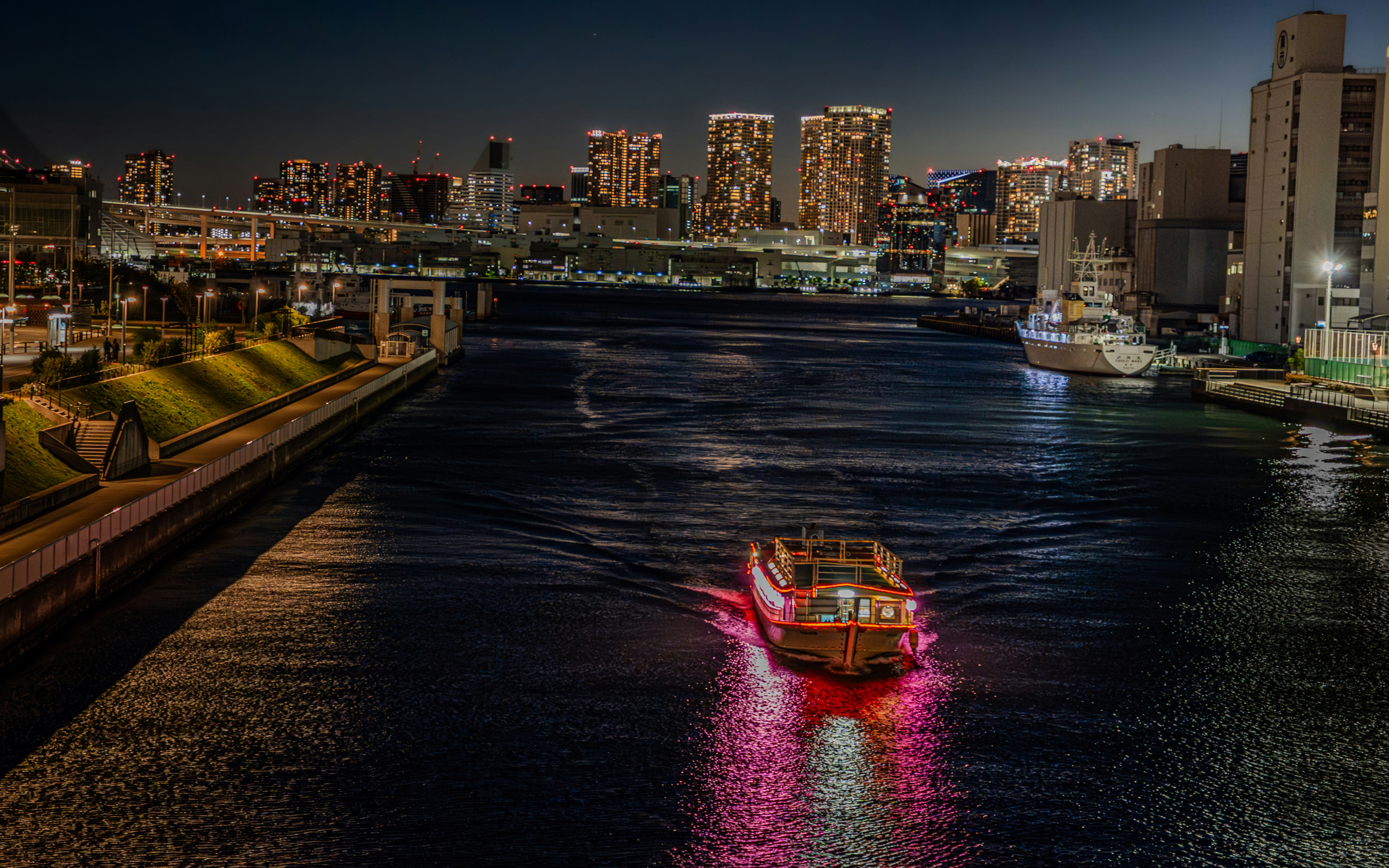 A beautifully composed night shot of Tokyo’s waterfront, capturing a glowing yakatabune (traditional Japanese pleasure boat) cruising through the river under the shimmering city skyline. The reflections of neon lights dance on the calm dark water, while the illuminated buildings and bridges in the background create a perfect contrast between Tokyo’s modern cityscape and its timeless charm. The soft gradient of the night sky adds a serene balance to the vivid urban lights.
