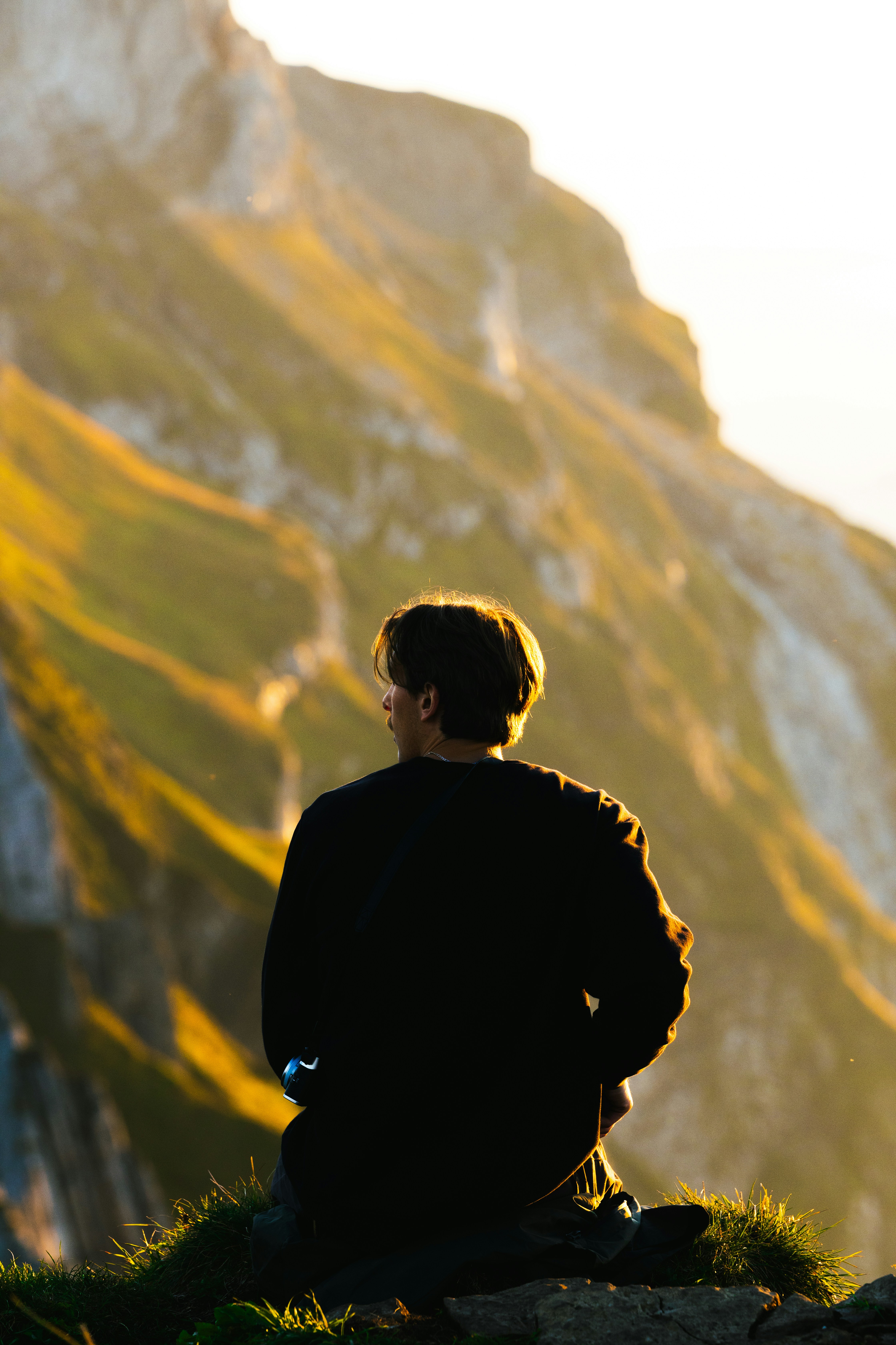 A man sits near the edge of a mountain cliff at golden hour, facing the glowing peaks of the Swiss Alps.