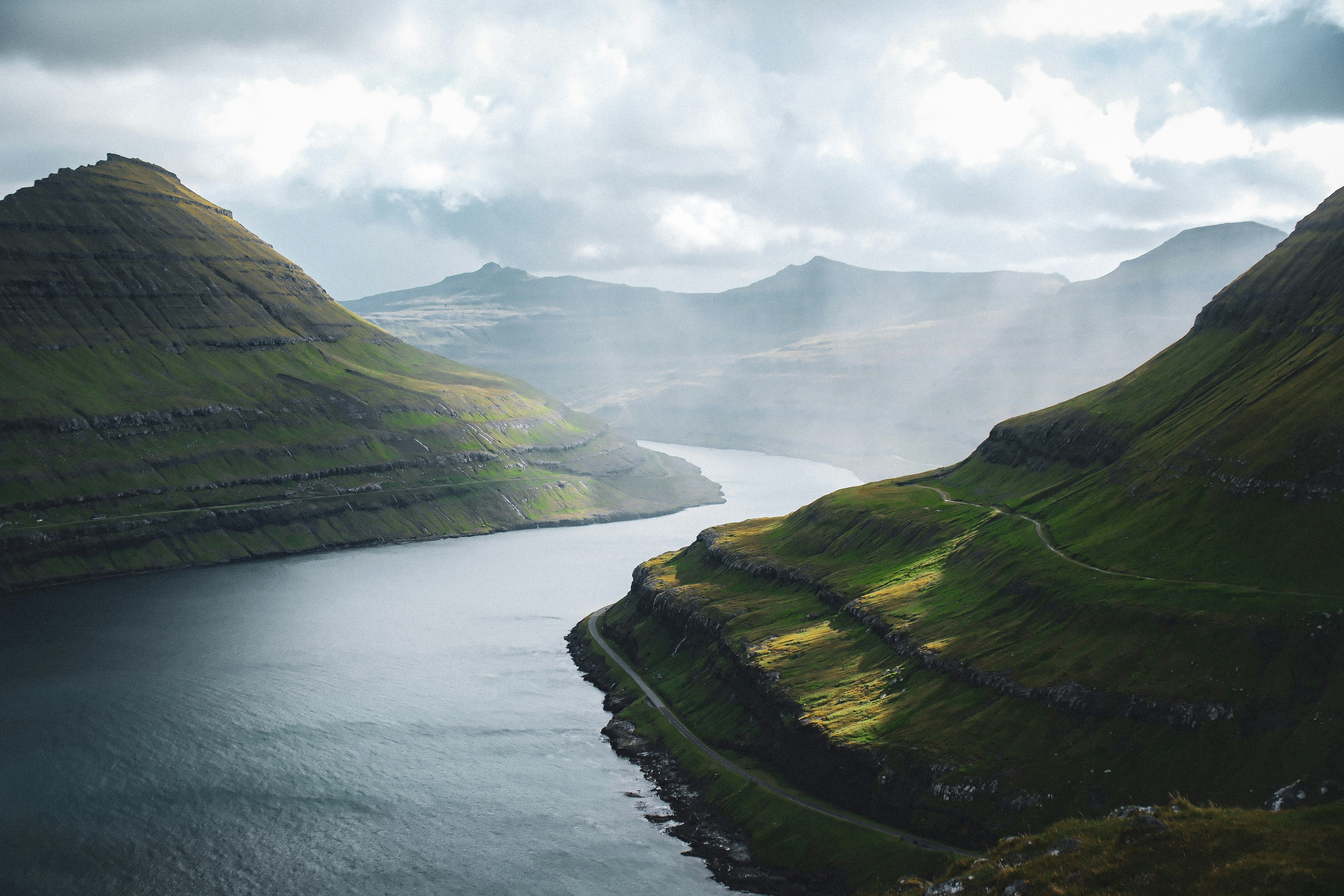 A winding fjord cuts through steep green mountains on the Faroe Islands, illuminated by sunlight breaking through the clouds.