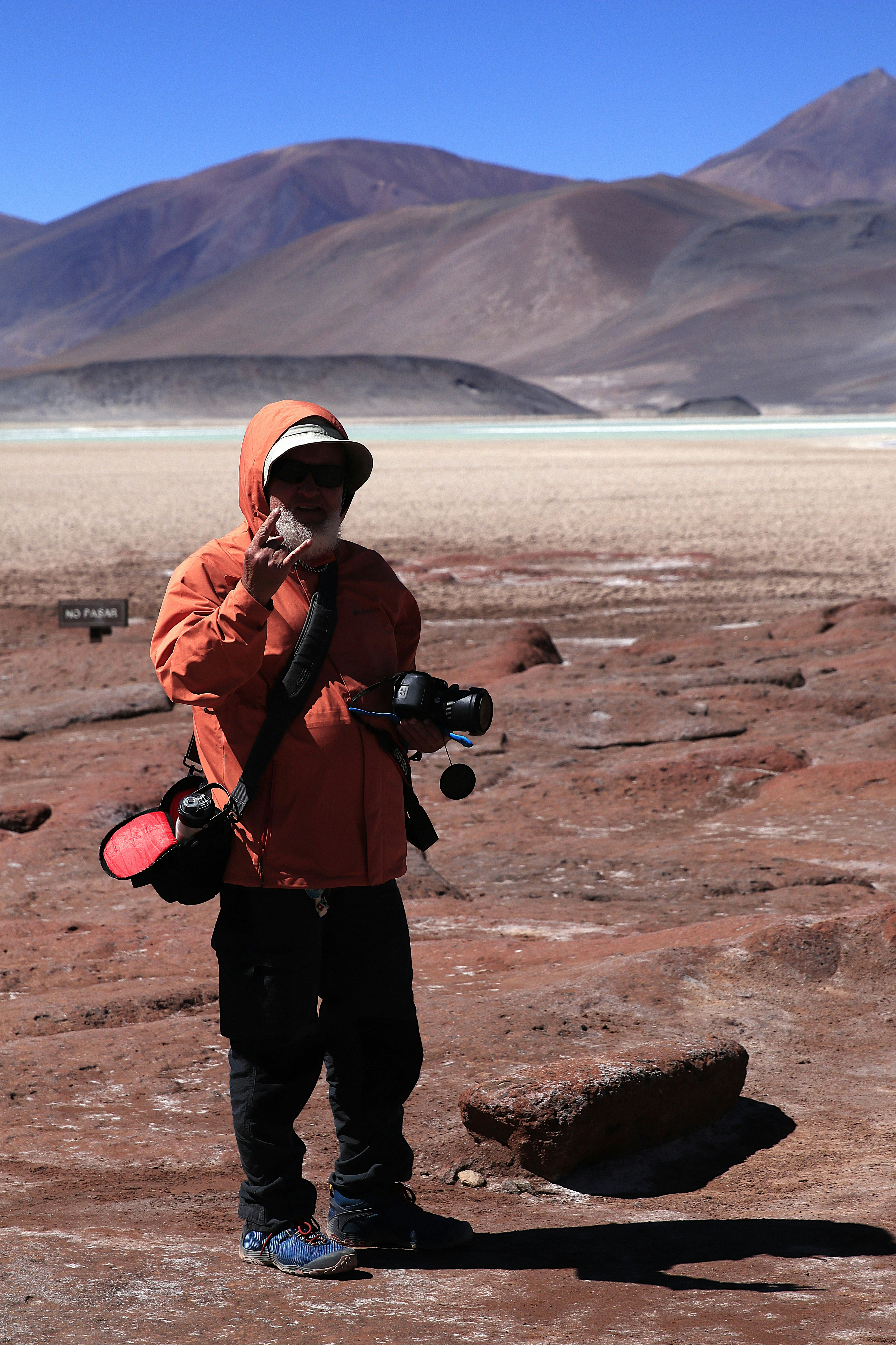 Red-Jacket Photographer at the Edge of a Turquoise Salt Lake Beneath a Pallid Mountain Range