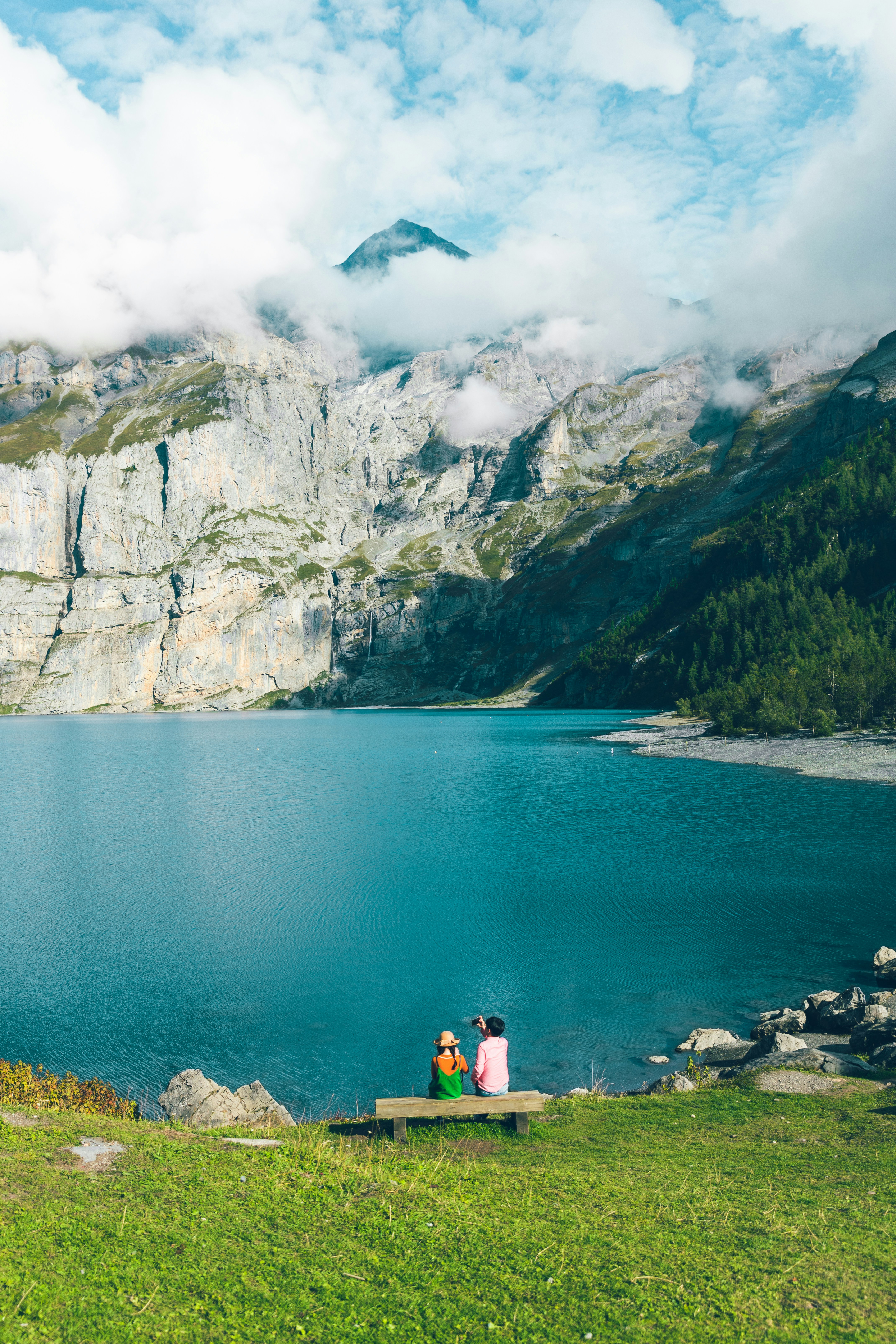 Two people sit on a bench near the shore of Oeschinensee. Behind them, turquoise water meets tall limestone cliffs partially covered with mist and scattered forest.