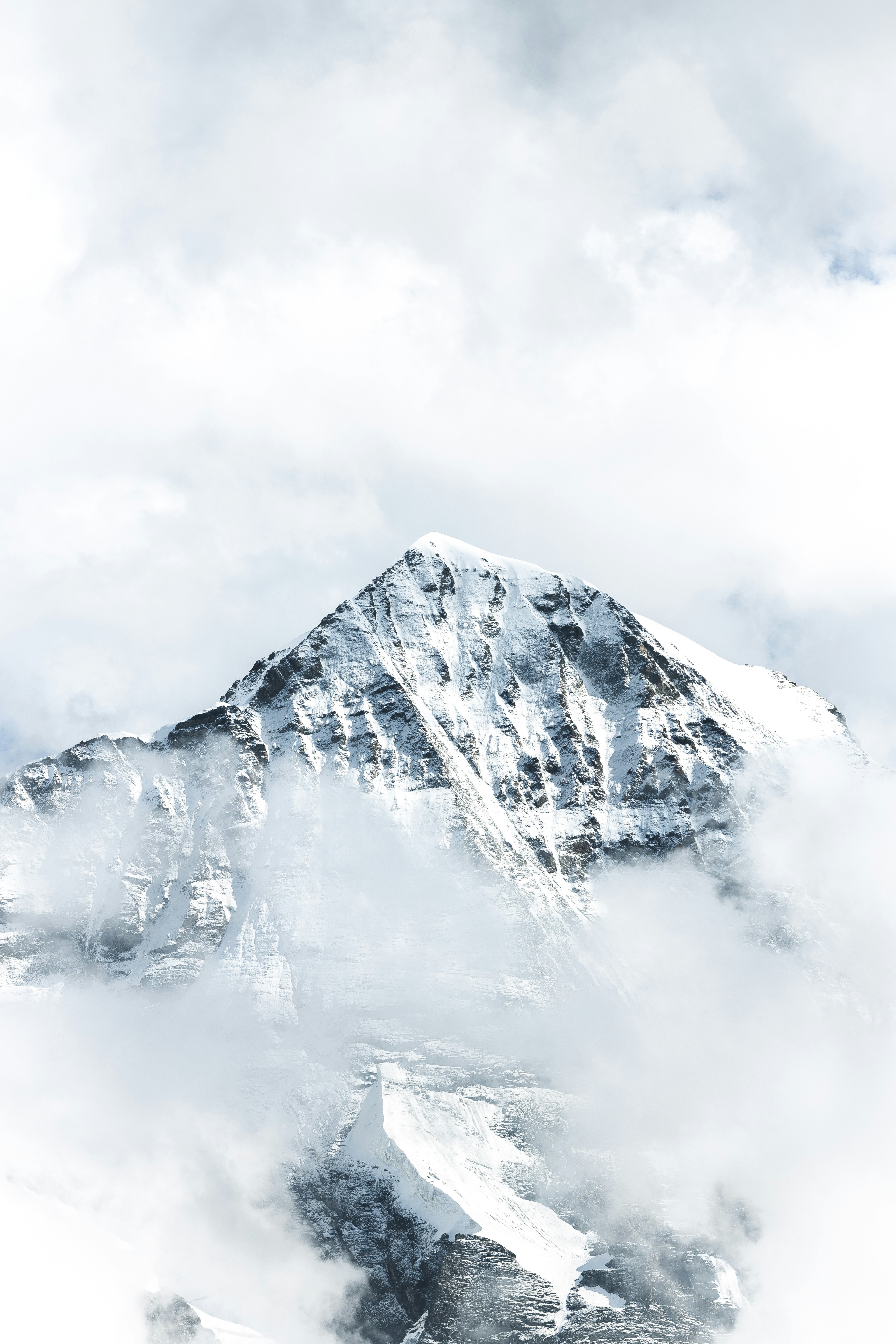 he snow-covered summit of the Eiger emerges through dense white clouds, its steep rock face faintly visible. The photograph captures the scale and isolation of the high Bernese Alps.