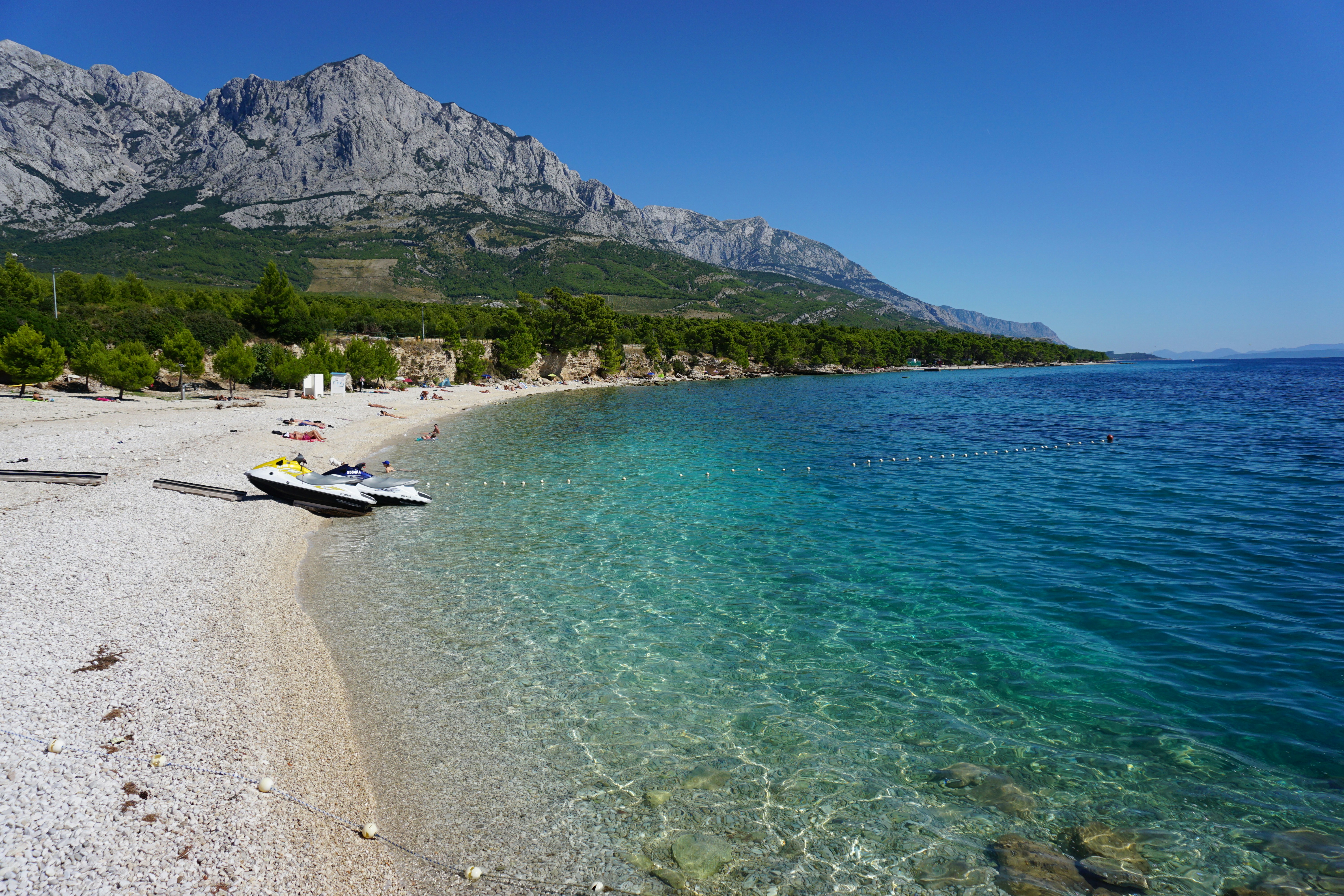 Sunlit Aegean Edge: Crystal Waters Meet Pebble Beach and Limestone Mountains