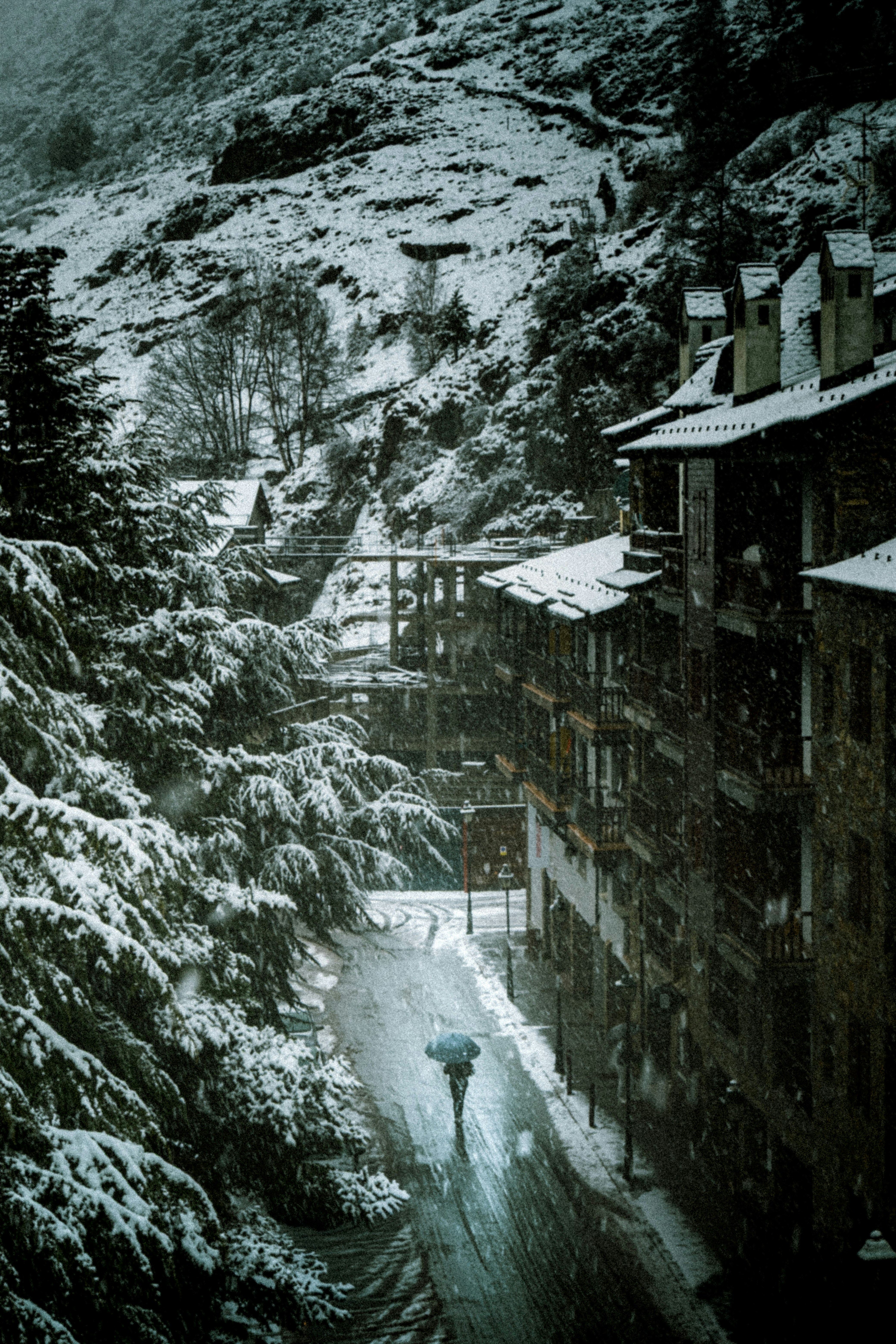 A person walks down the street in Andorra La Vella, during winter, while it's snowing. The roads and mountains are filled with snow, and the light was almost gone, giving it a more gloomy look.