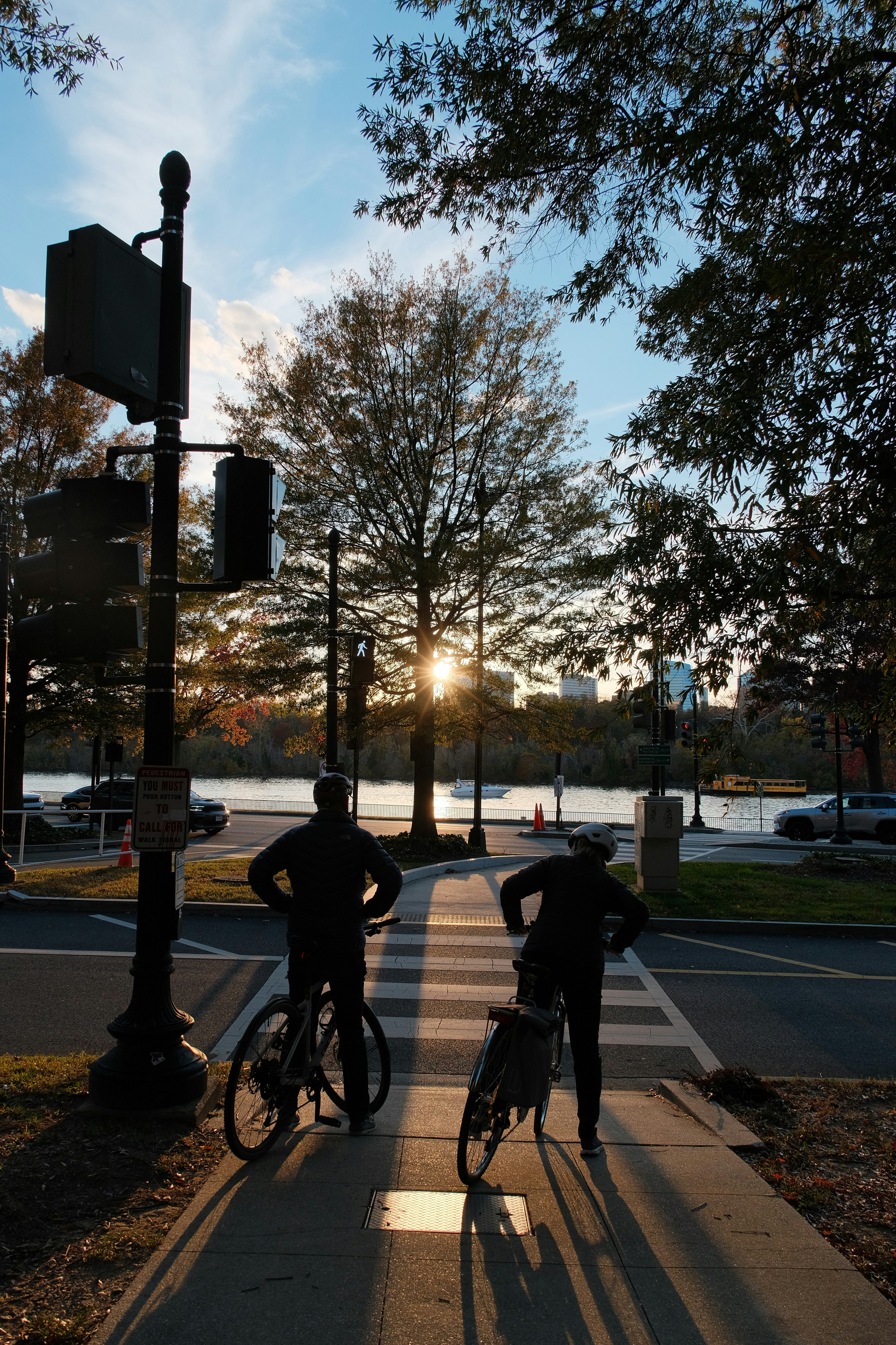 Sunlit Standstill at Riverside Crosswalk: Two Cyclists in Dappled Evening Light