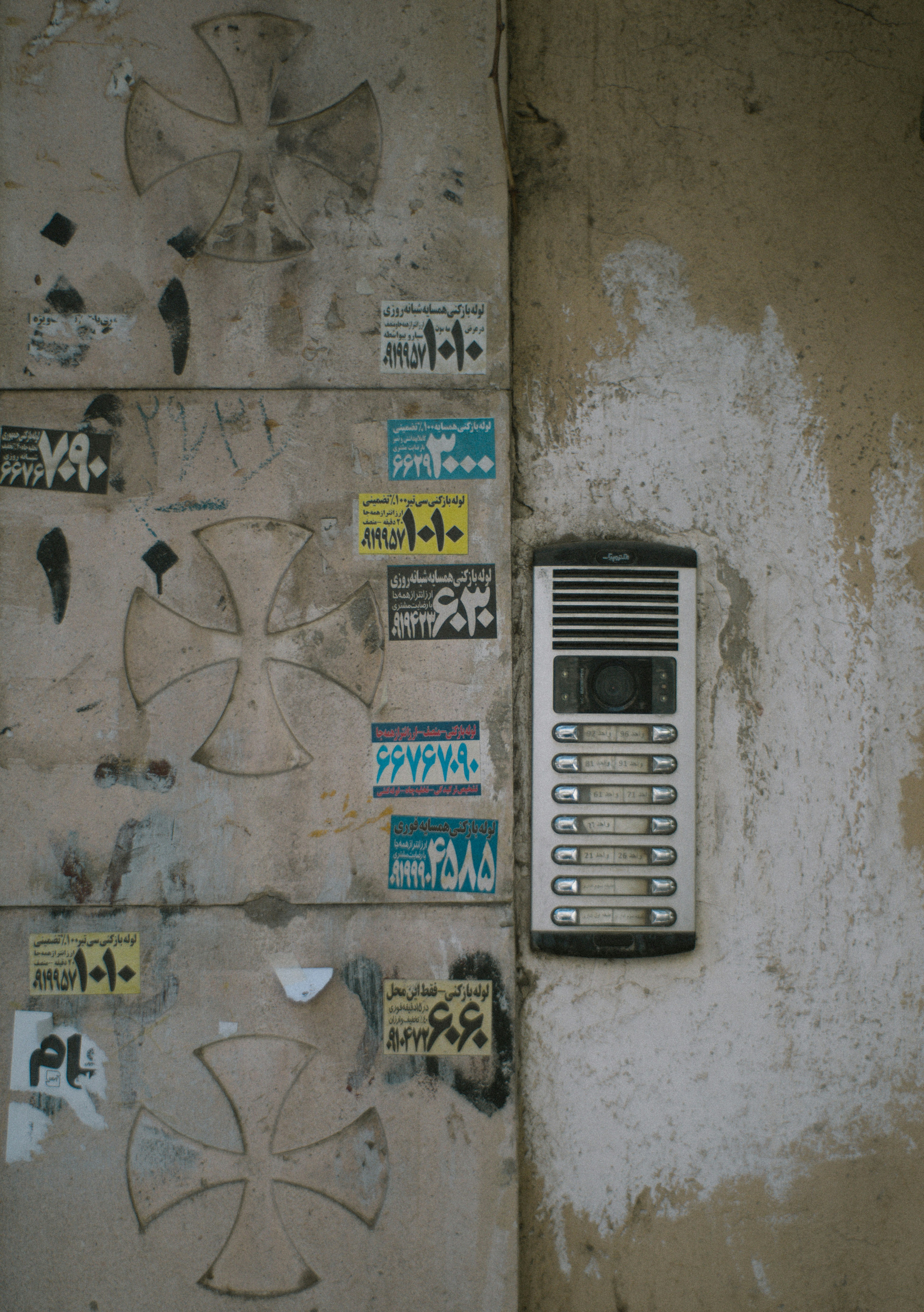 Weathered concrete wall features a metal intercom panel flanked by faded emblems and a collage of stickers. Colorful tags add urban texture, hinting at lived-in history.