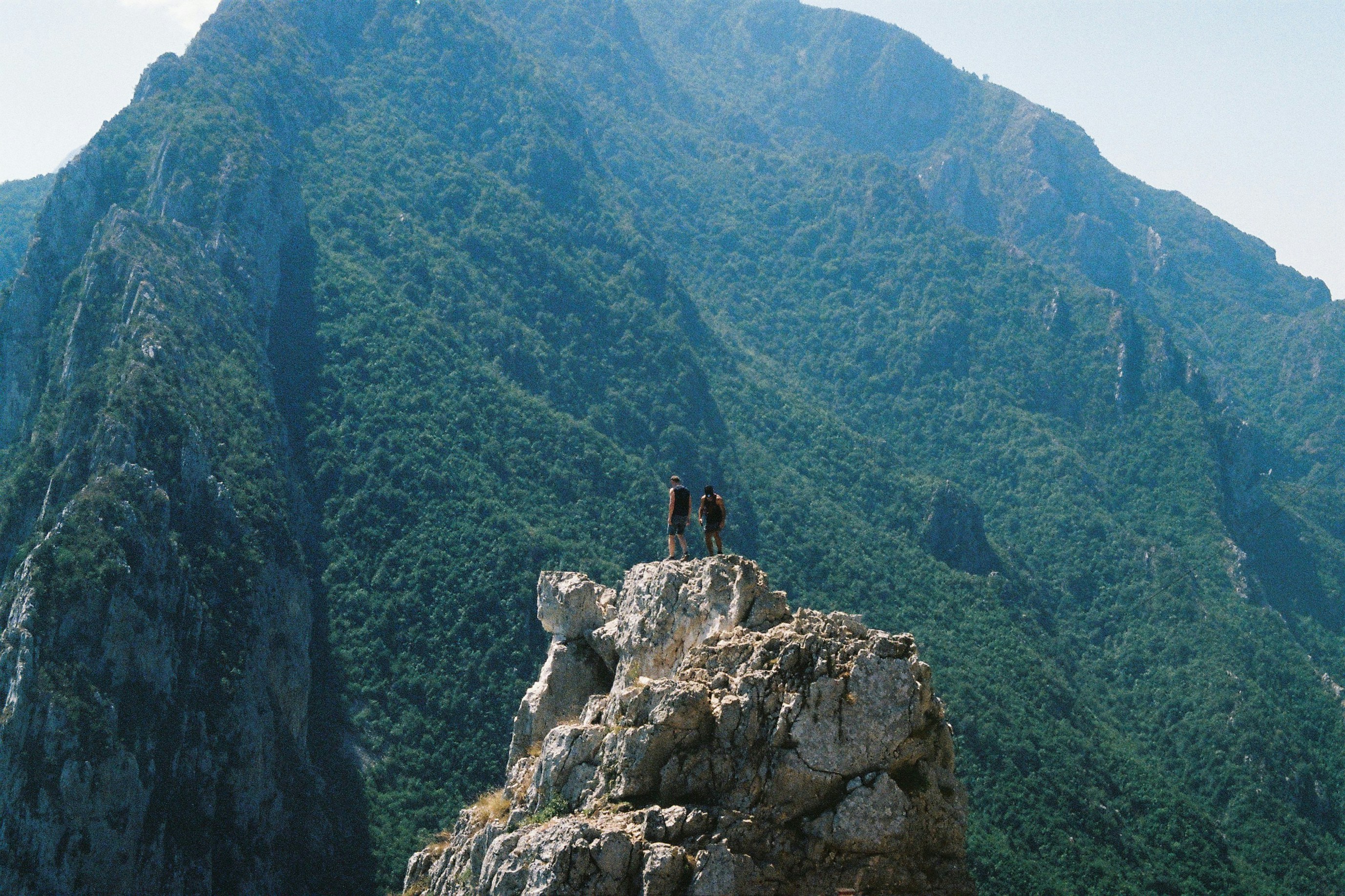 Two hikers stand atop a rocky peak with mountains behind.